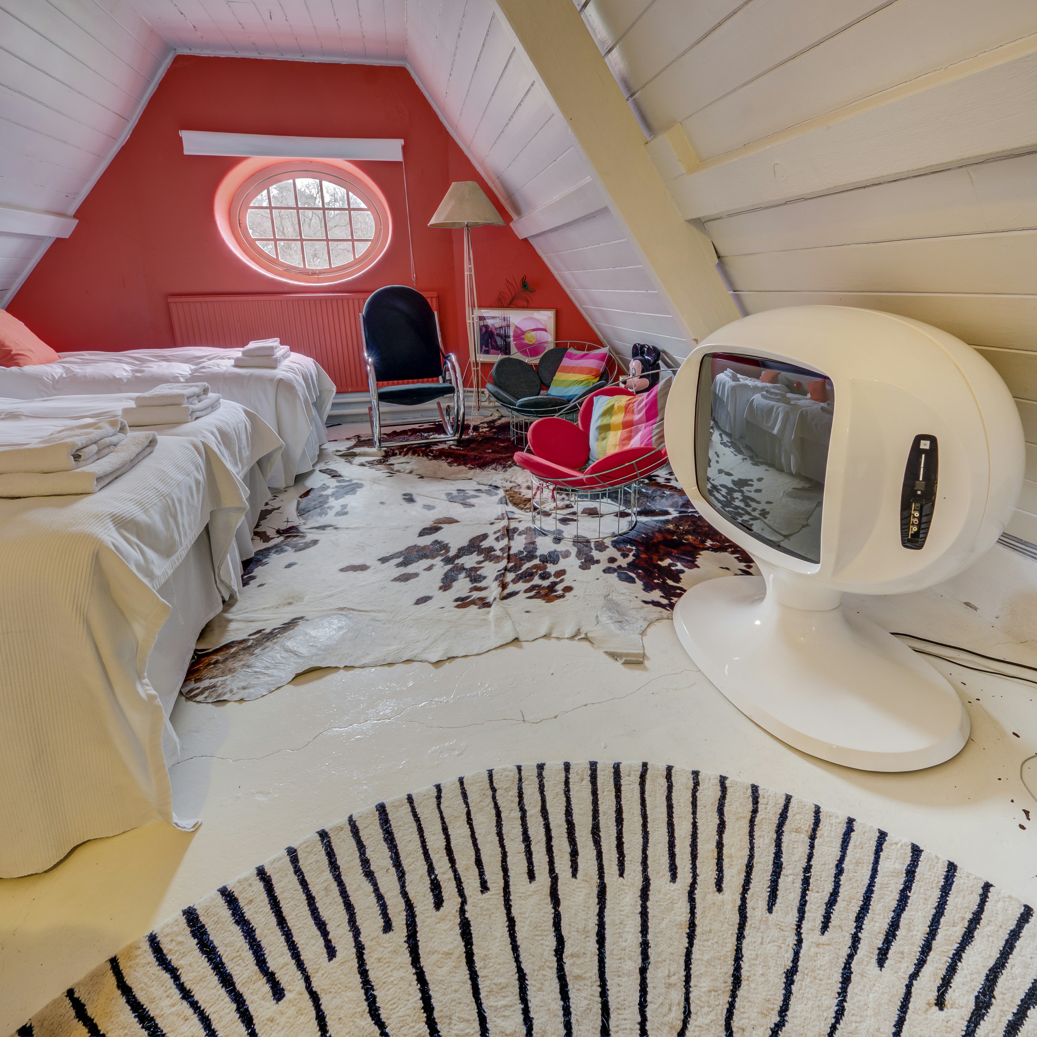 A quirky attic bedroom with a curved, retro TV and bright accents sits beneath a pink circular window; white beds line one wall while an eclectic rug and colorful chairs add playful character.