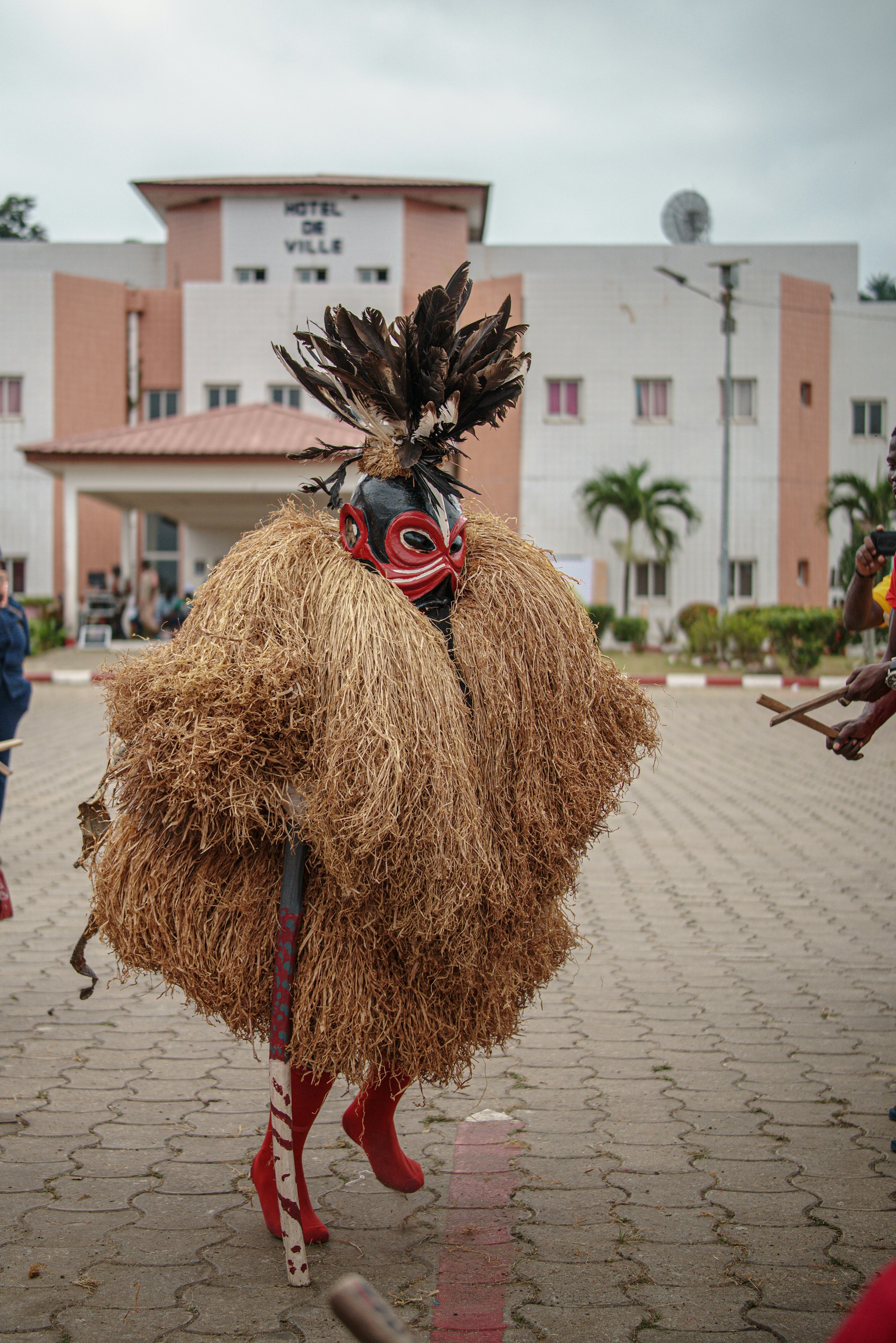 A performer in a straw costume and red mask strides across a courtyard, with a pastel building backdrop and onlookers nearby. The scene captures a festive street performance.