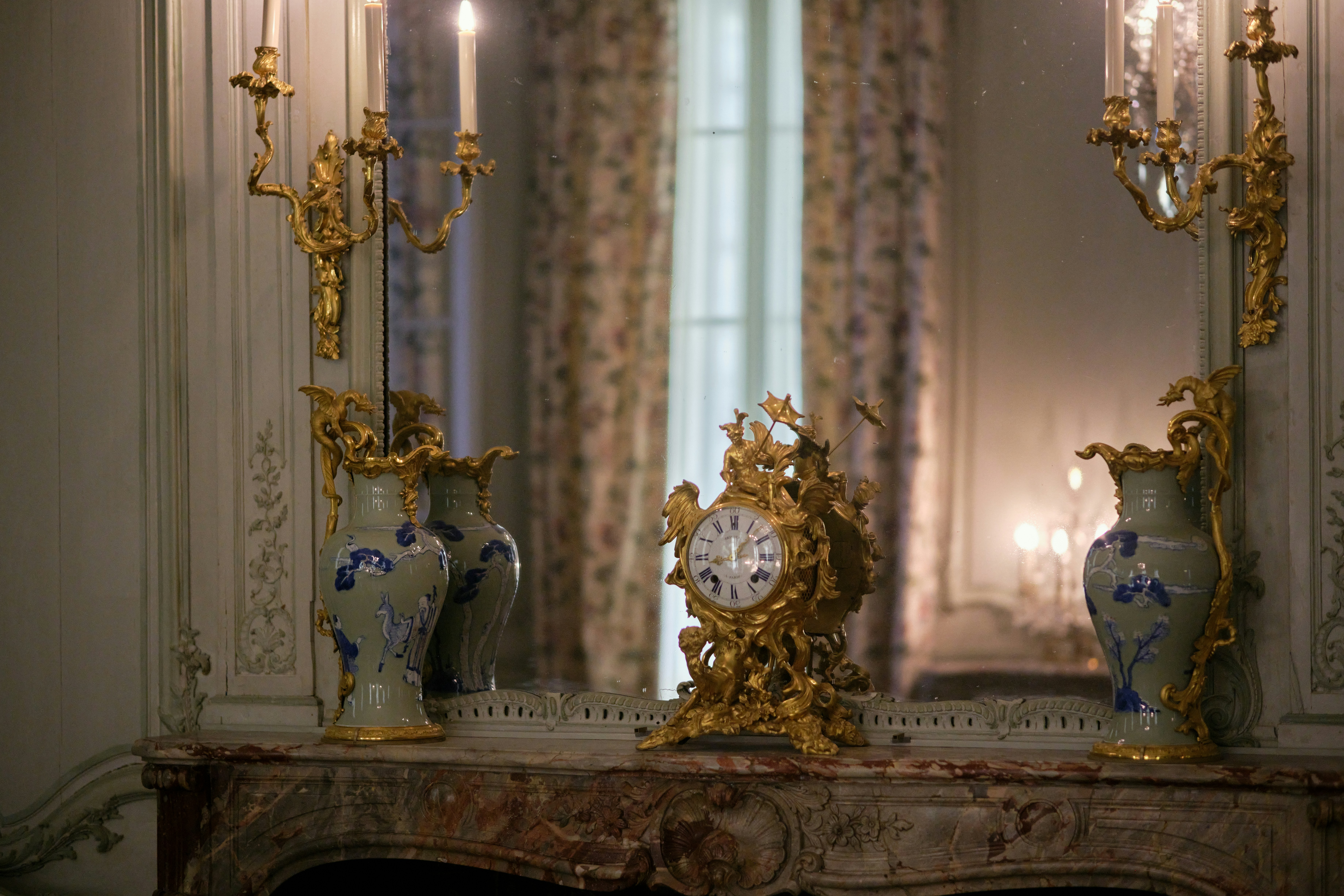Luxurious mantel with a gilded clock flanked by porcelain vases and candelabras, reflected in a dimly lit mirror-backed room.