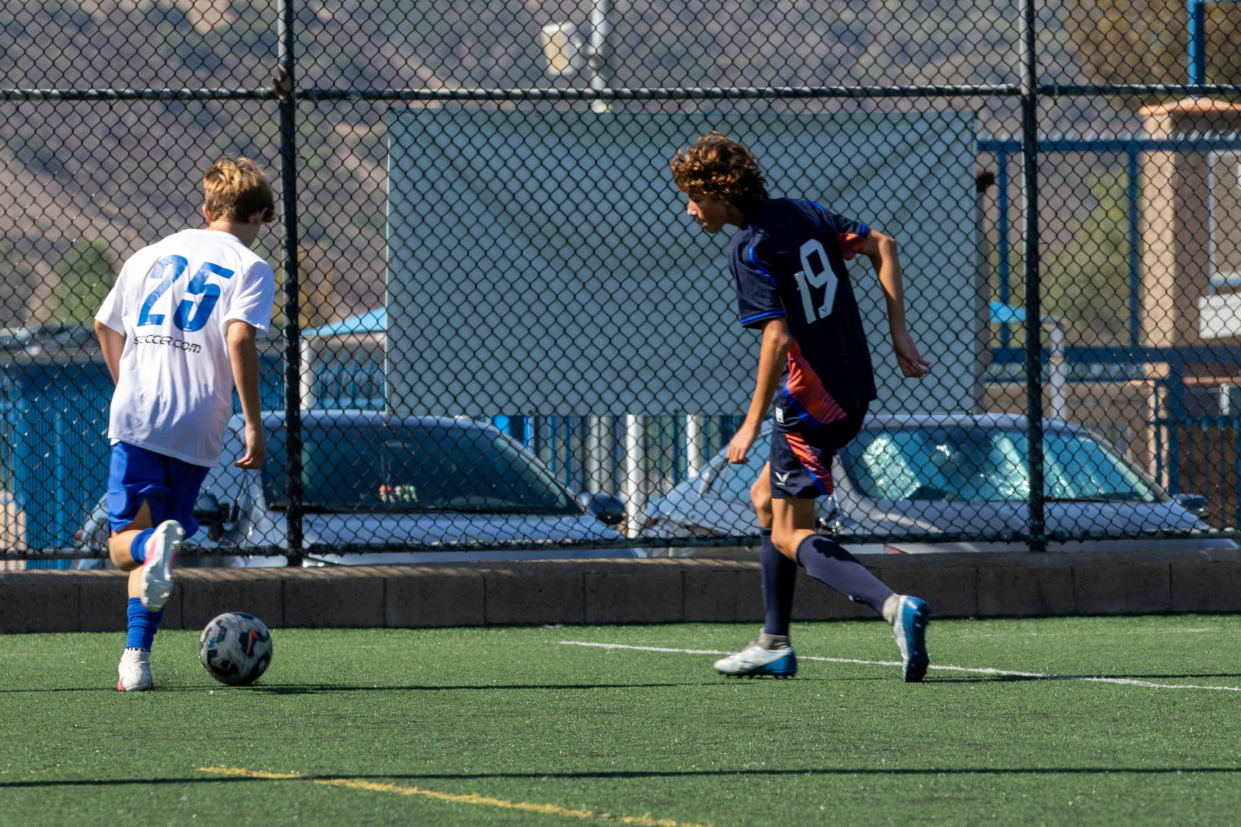 Midfield duel: two youth players chase the ball under a chain-link backdrop