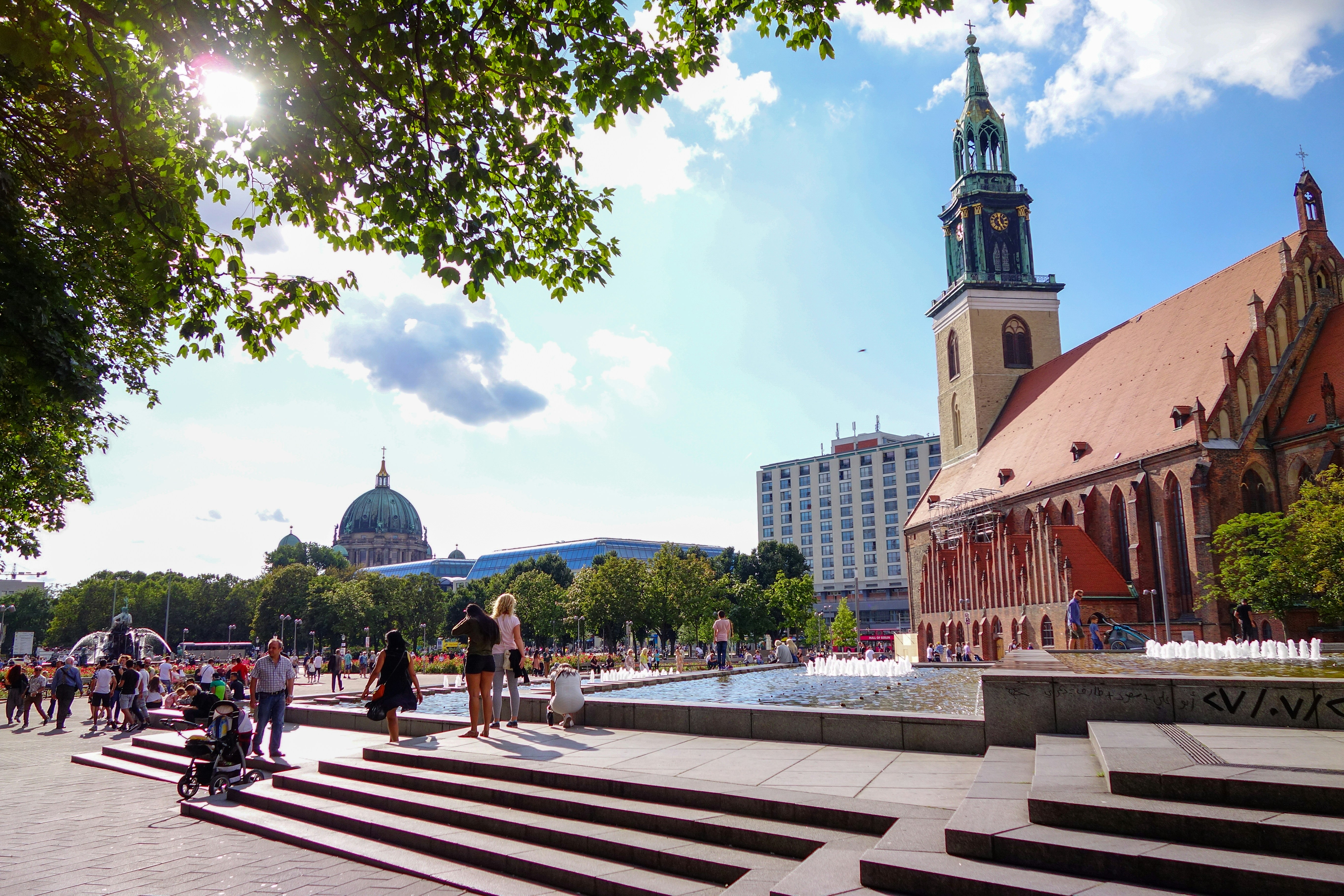 The Gothic spire of St. Mary's Church rises above a fountain plaza in central Berlin, Germany, with the Berlin Cathedral dome visible in the distance under a partly cloudy sky.