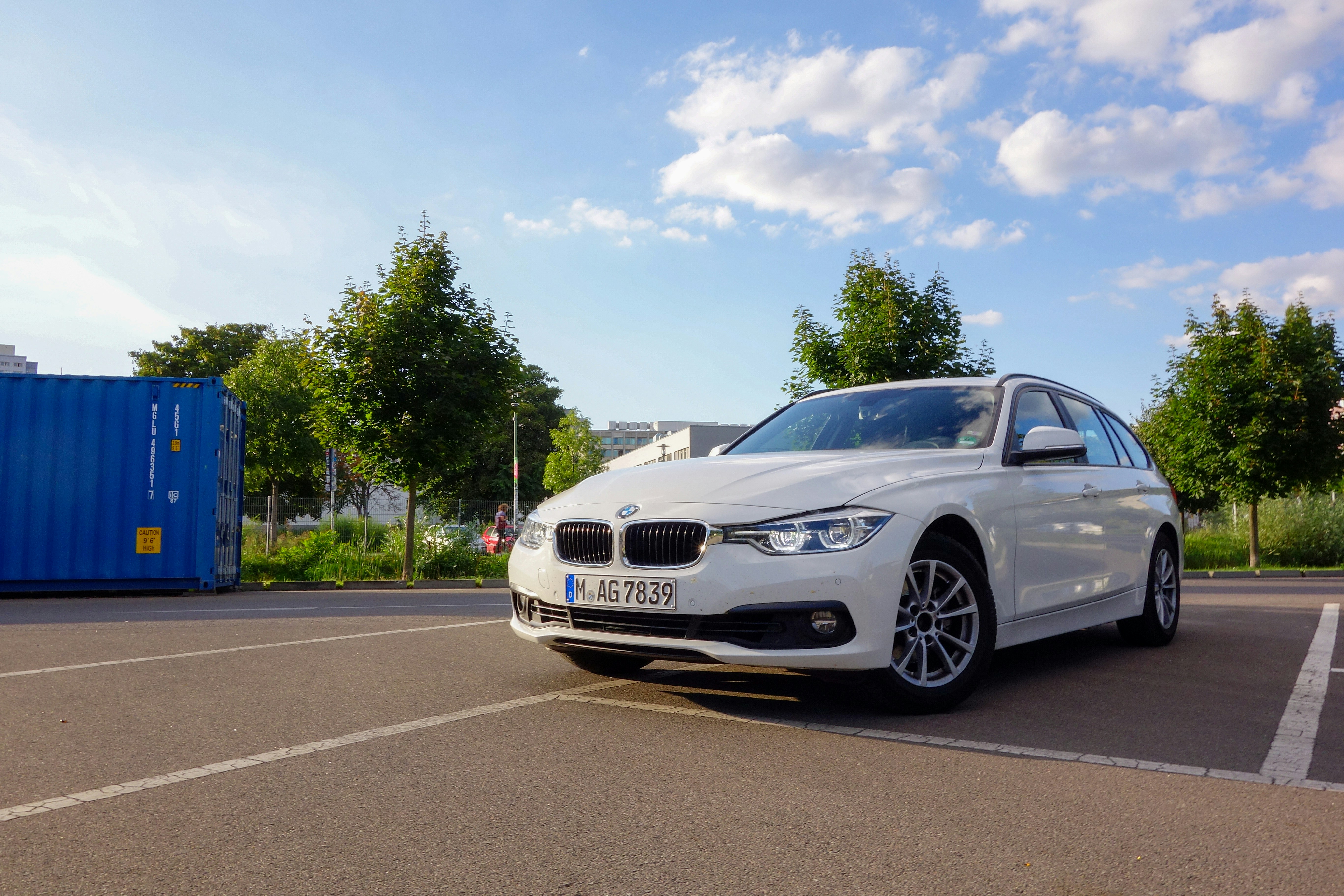 White BMW in a Sunny Parking Lot with Greenery and Delivery Container