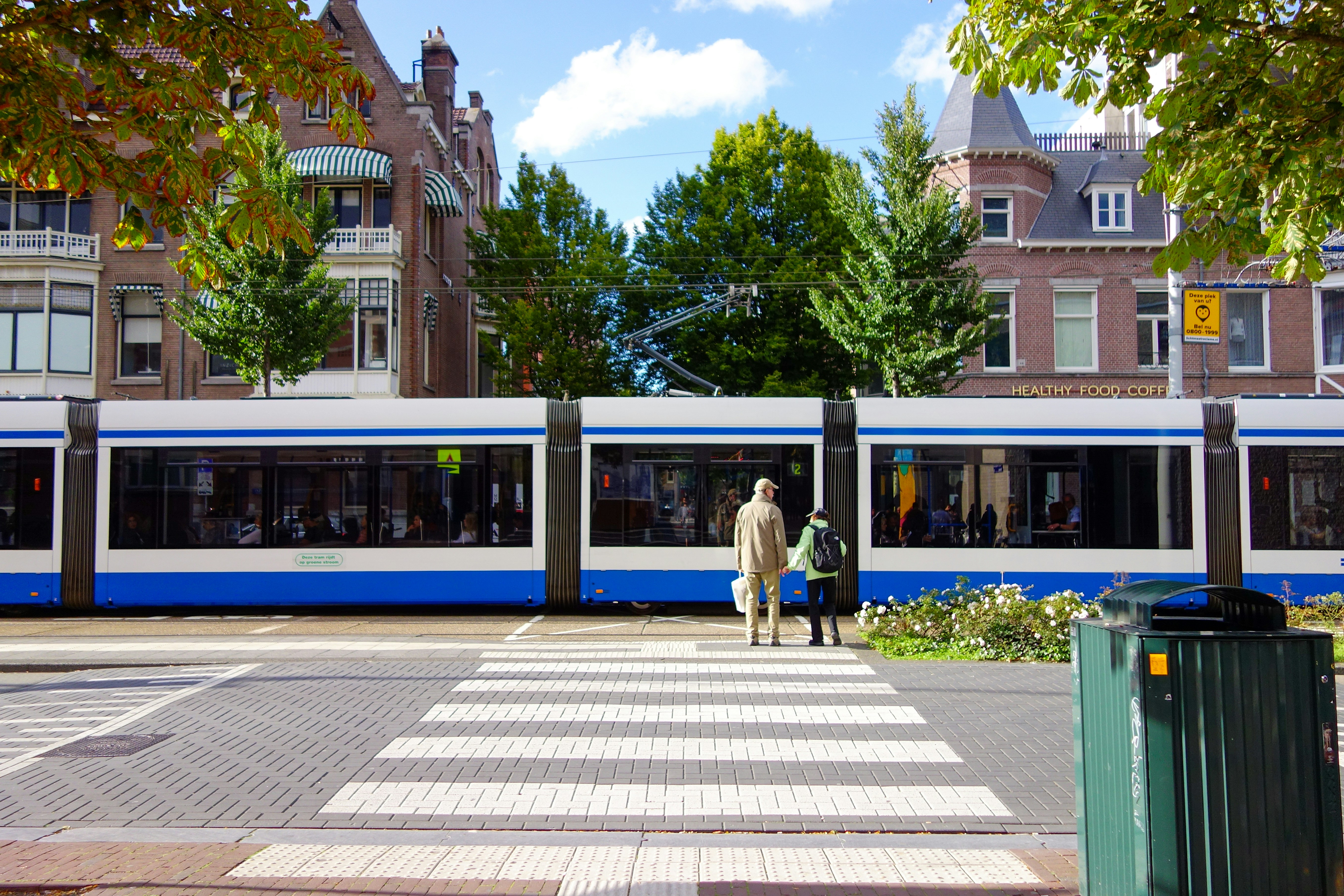 Two passengers wait to board a blue-and-white tram across a sunlit pedestrian crossing
