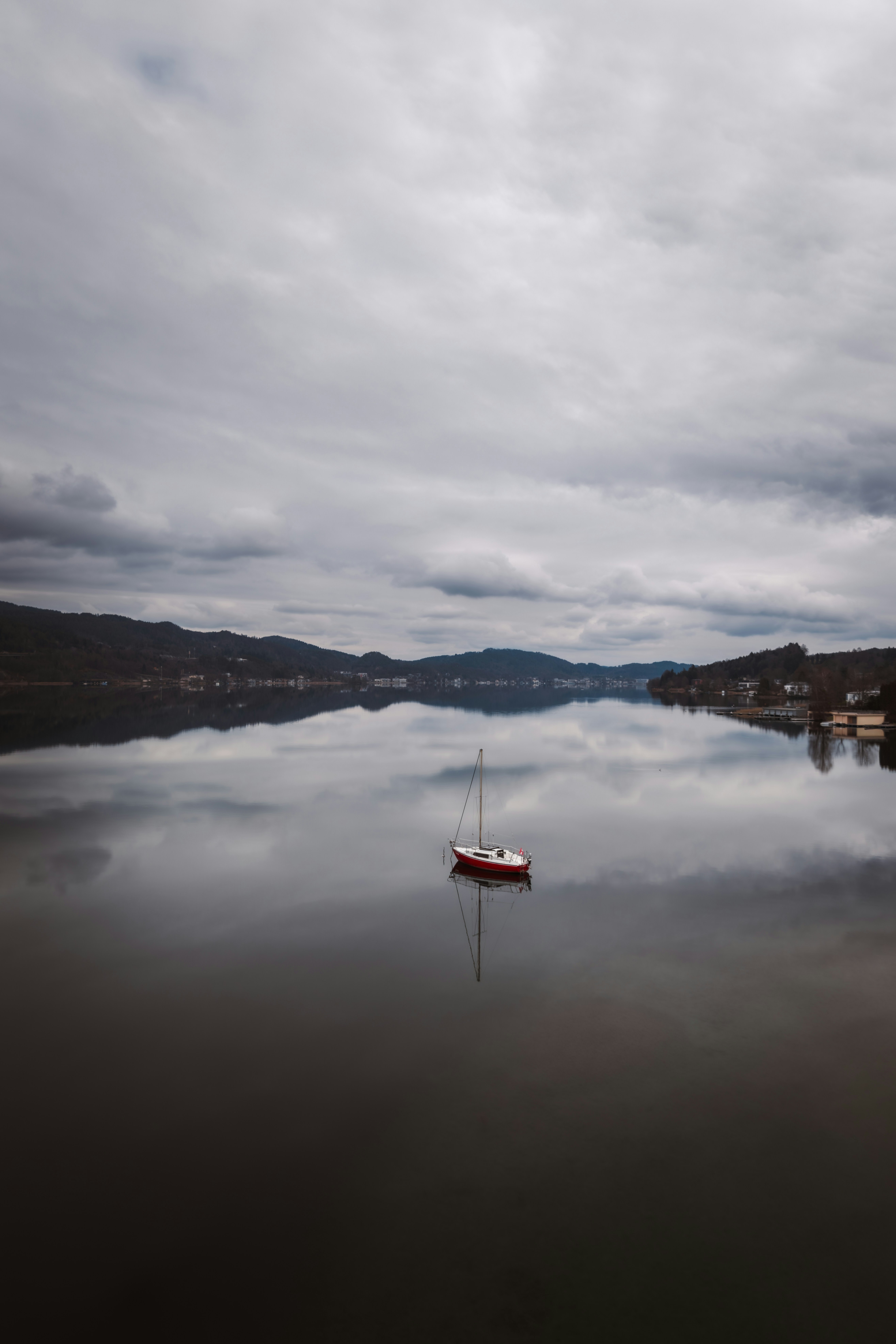 Solitary red sailboat on a mirrored fjord under a clouded sky