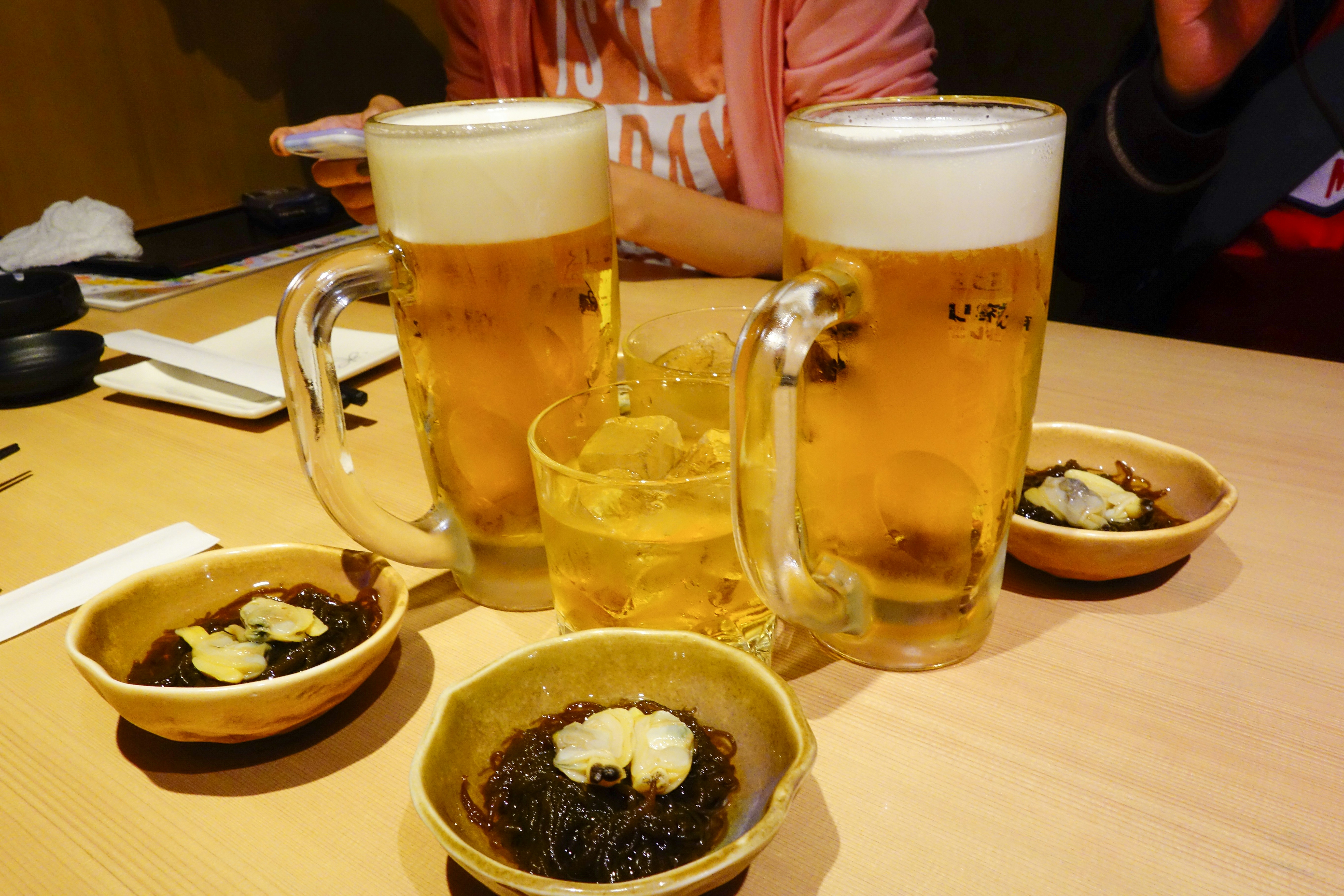 Two frosty mugs of draft beer and iced oolong tea on a wooden table beside small dishes of seasoned oyster with fermented sauce at a Japanese izakaya.