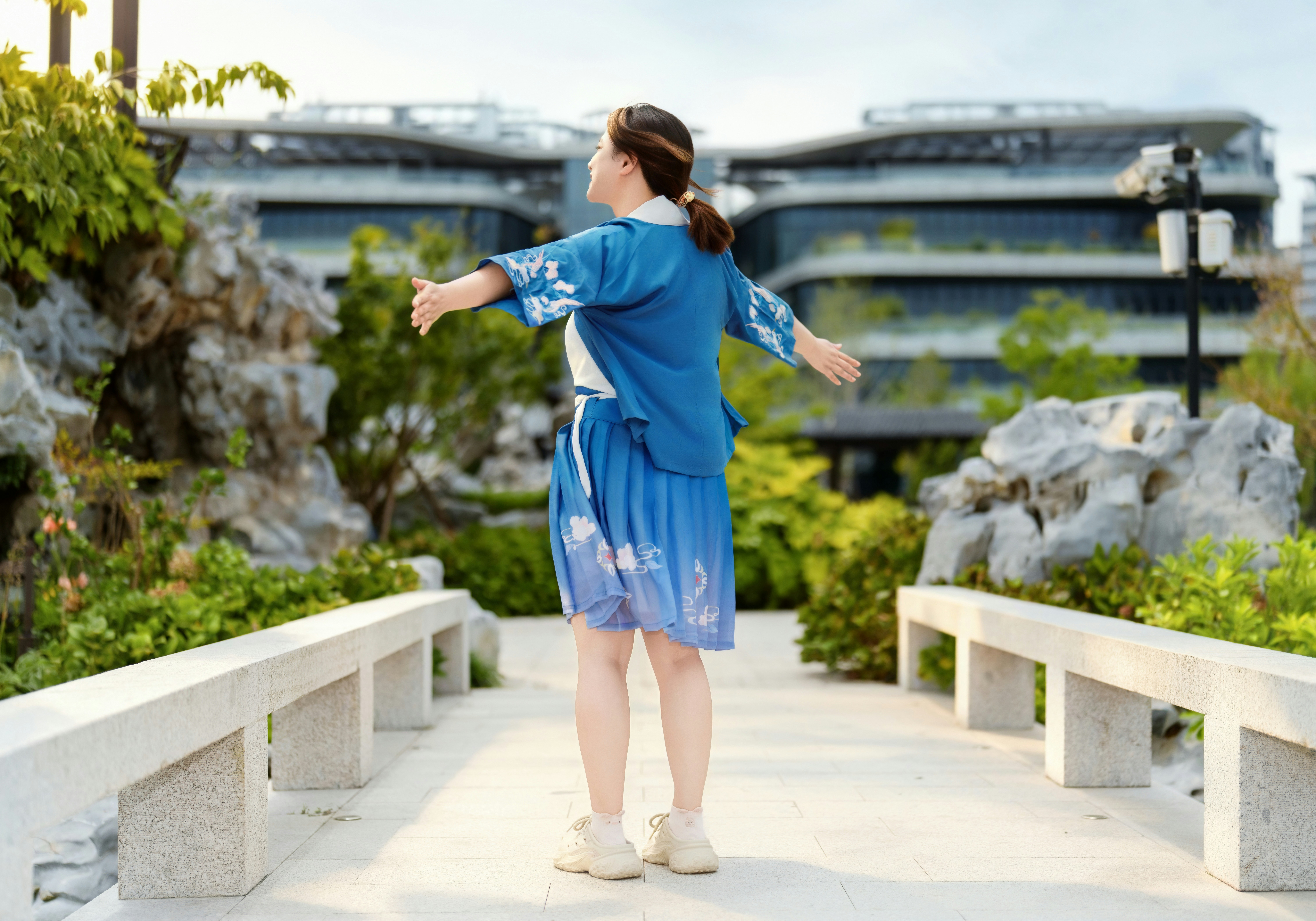A woman in a blue kimono-style dress stands with arms outstretched on a sunlit walkway amid rocks and greenery, with contemporary architecture in the background.