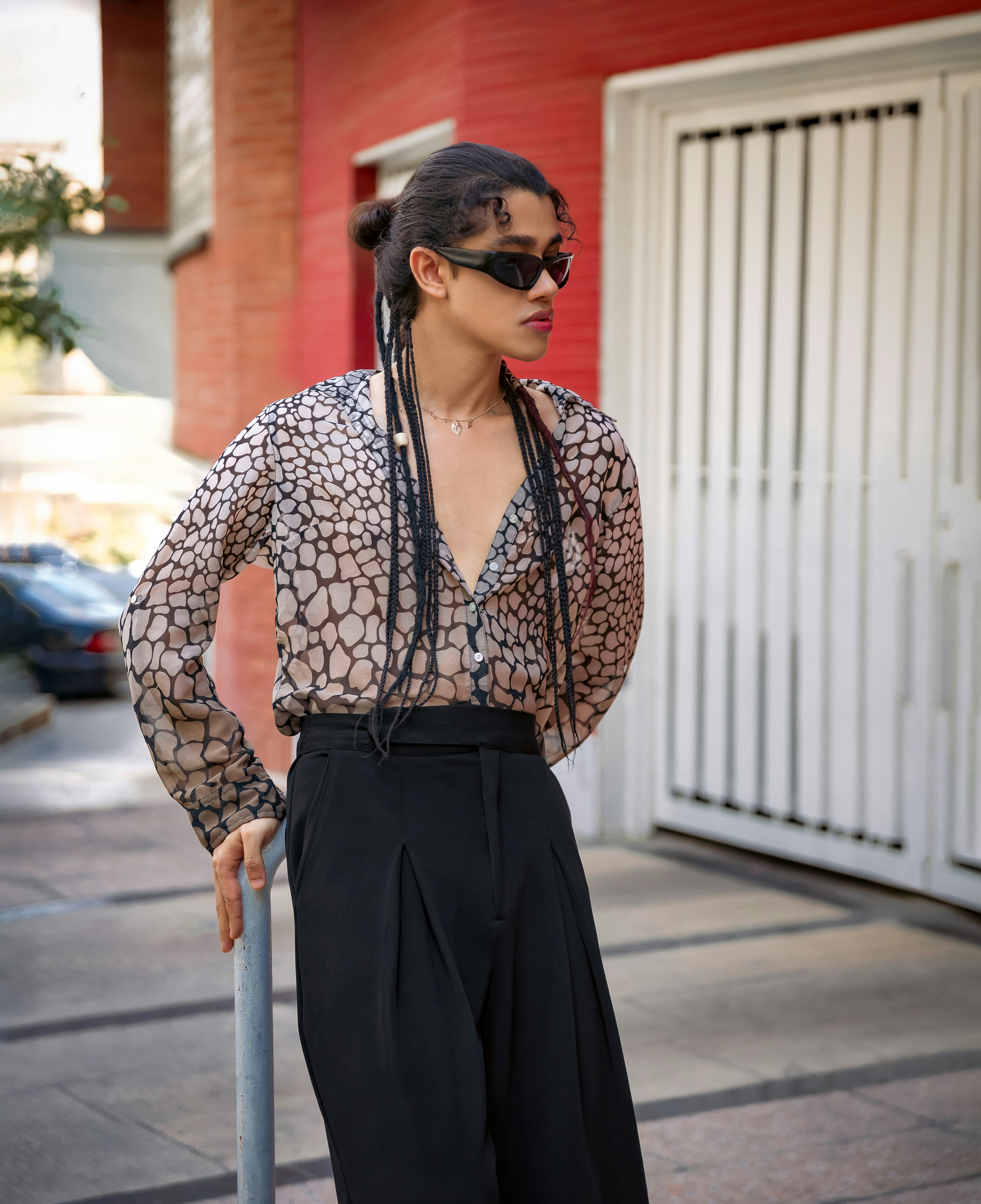 Woman with patterned sheer blouse and black high-waisted pants leans on a railing in an urban setting; angular sunglasses and braided hair create a strong, modern silhouette.