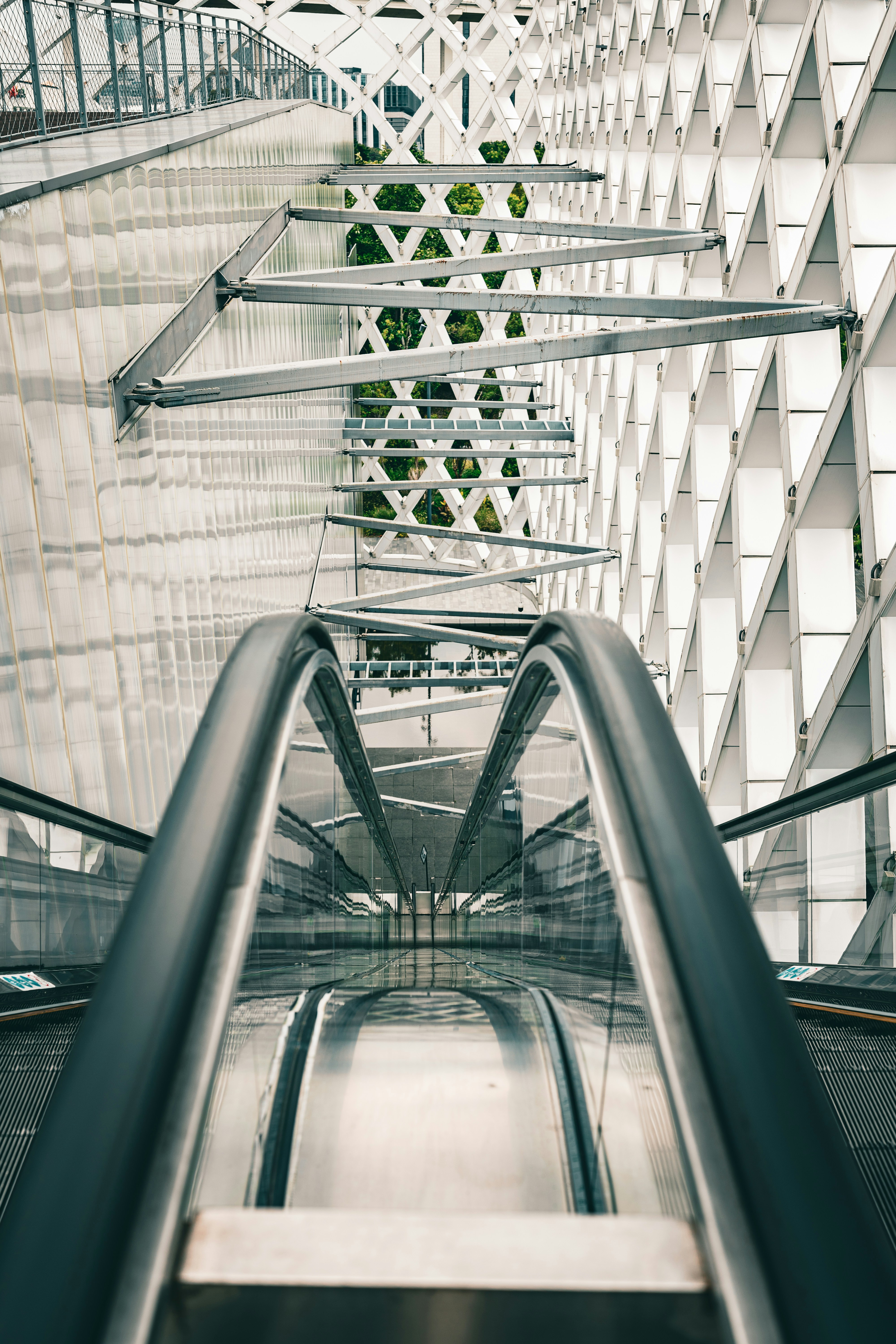 Escalators descend through a glass-walled, lattice-patterned atrium, capturing industrial geometry and reflections with muted tones. The scene emphasizes modern architecture and controlled movement. (Logo-free interior shot).