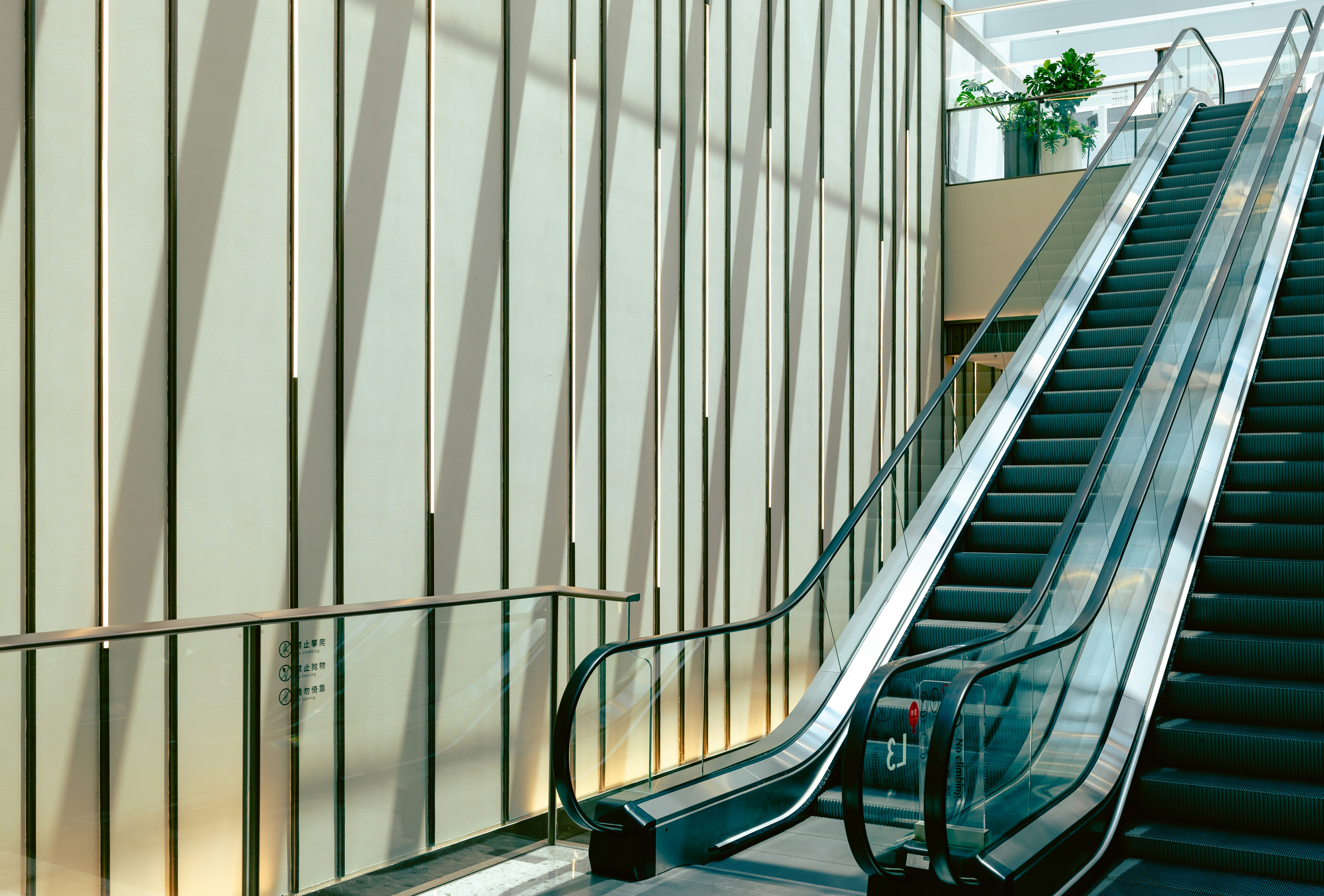 Escalators ascend beside a sunlit paneled atrium with vertical slats and a potted plant visible at the top. The scene emphasizes architectural geometry and scale.