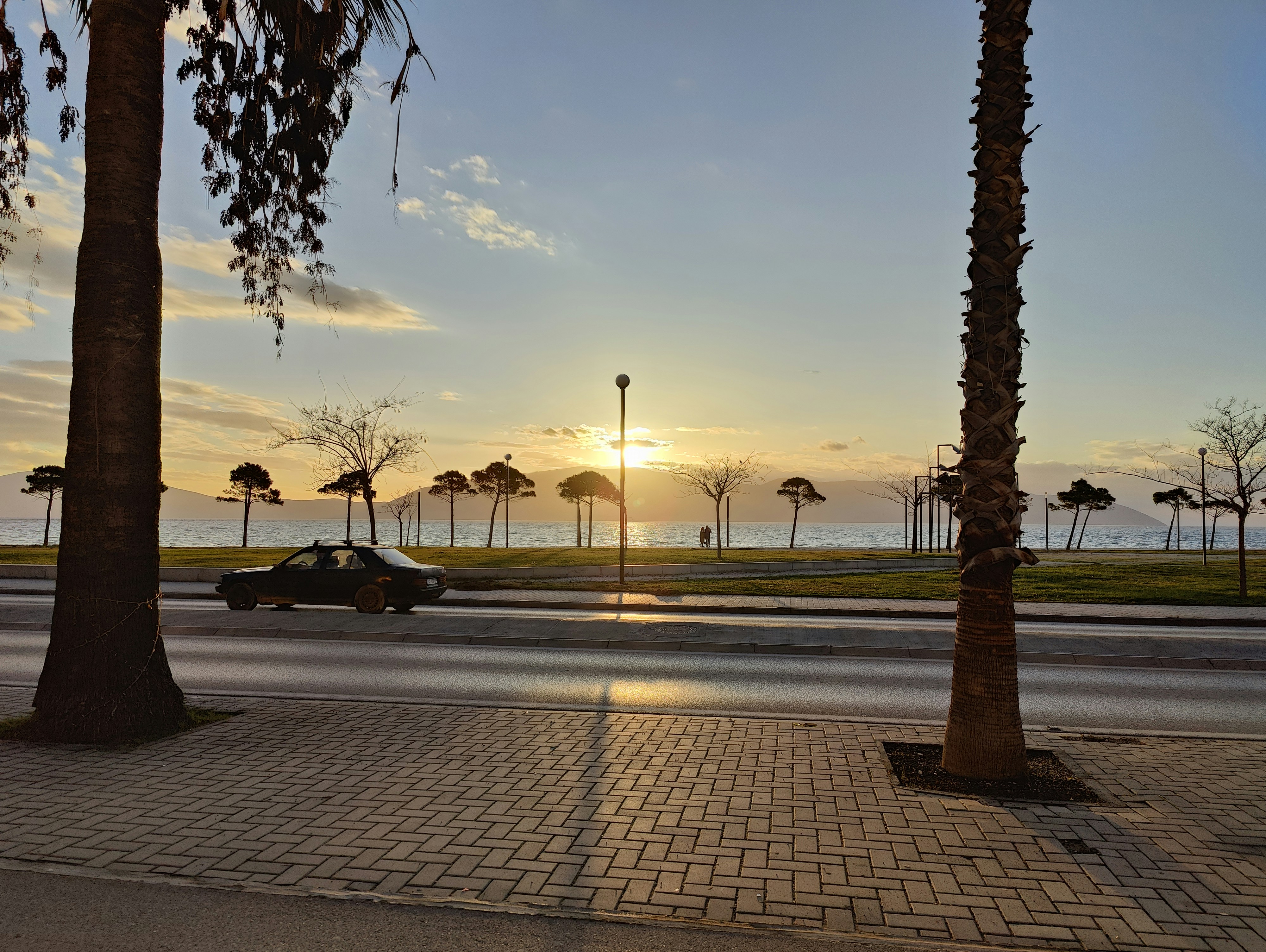 A tranquil coastal promenade at sunset, featuring tall palm trees framing a calm sea and a golden sun dipping below the horizon behind mountain silhouettes. A lonely car glides down the seaside road, bathed in warm, soft evening light