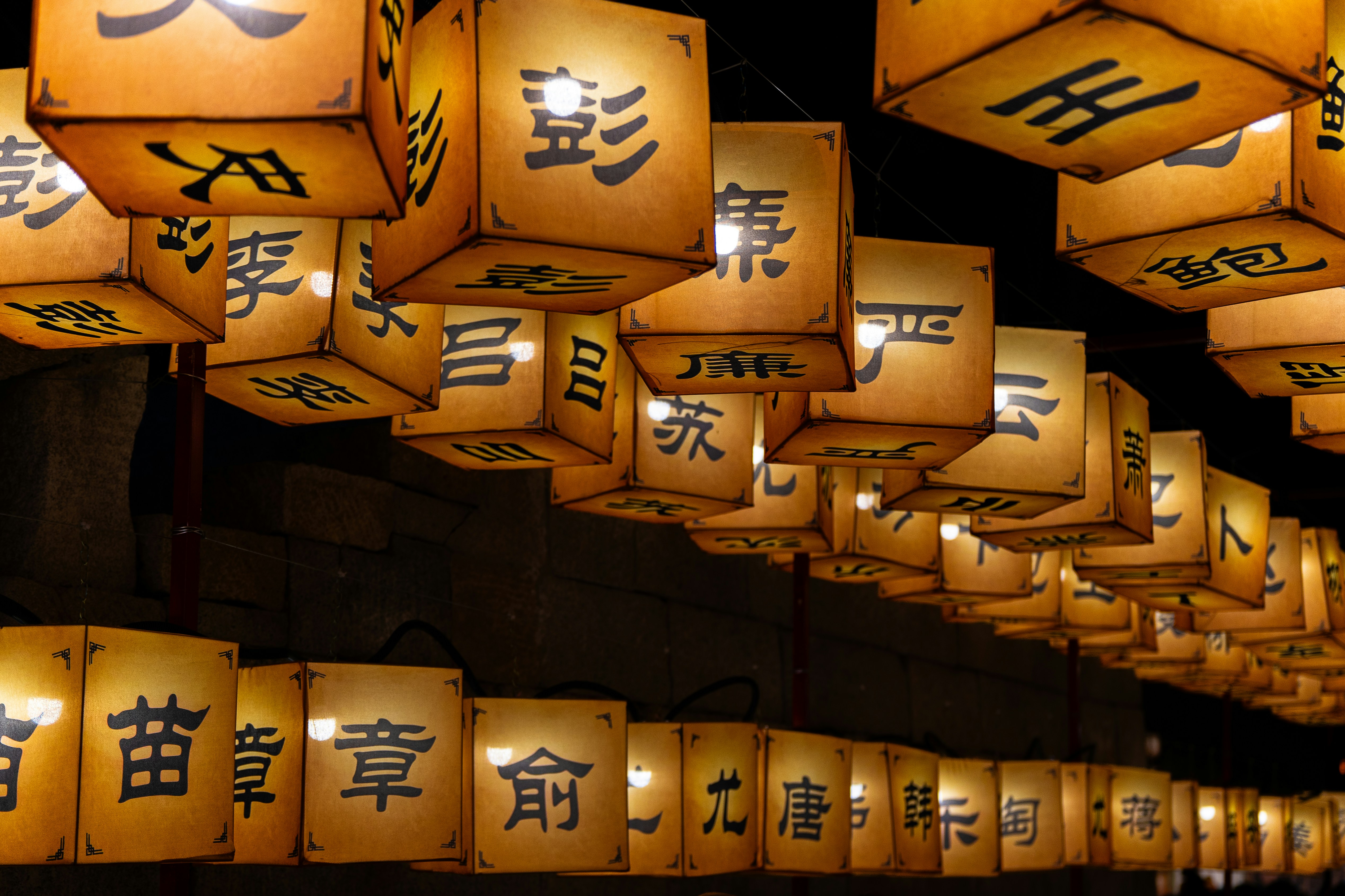 Rows of amber lanterns illuminate a dark wall, each bearing bold characters in a rhythmic, skyline-like arrangement; the shot captures a low-angle corridor of hanging lamps, suggesting a festive street scene.