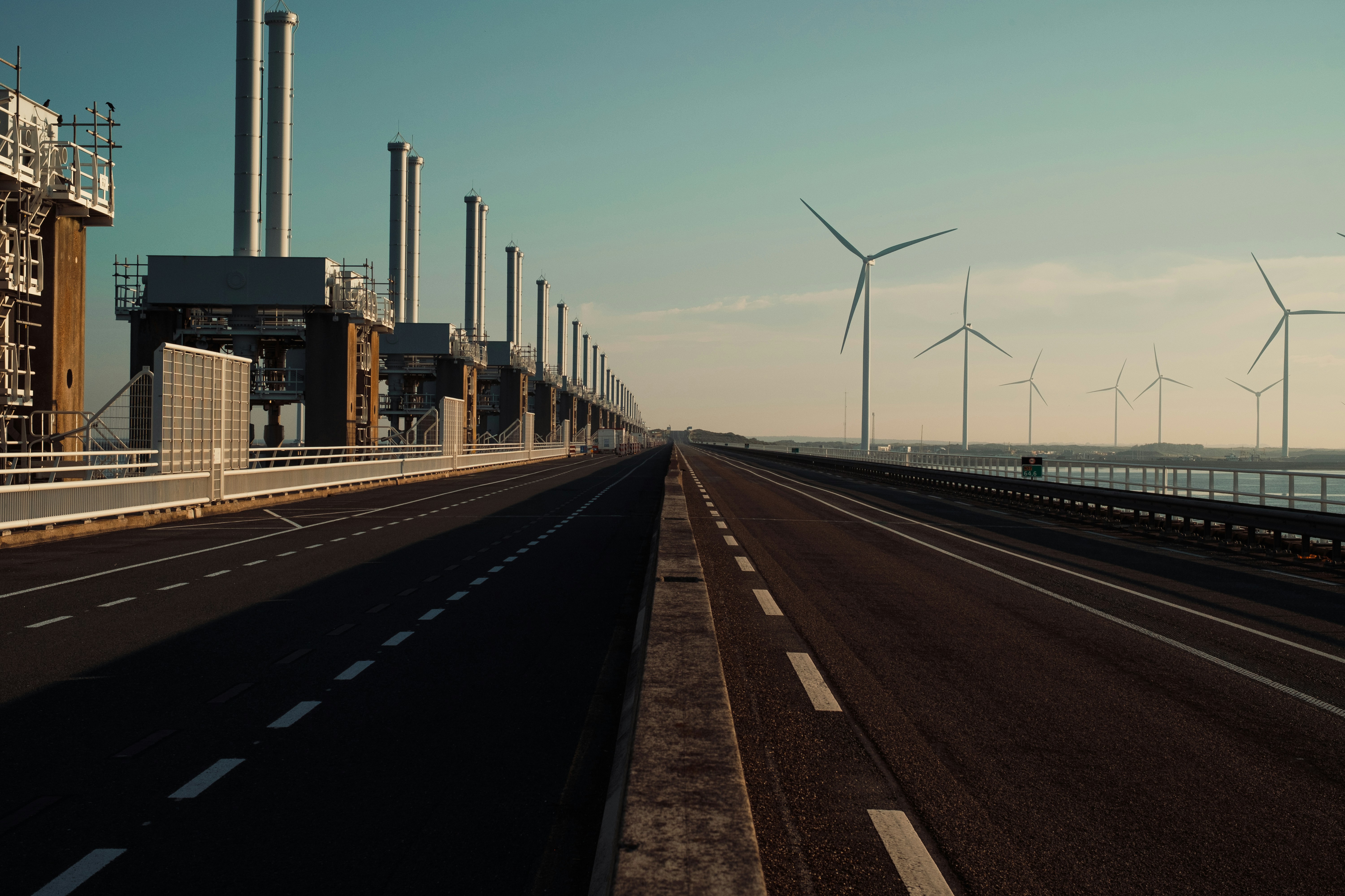 A serene, empty road stretching across the Oosterscheldekering storm surge barrier toward a distant vanishing point. Bathed in soft morning light, the central perspective highlights the massive industrial gate pillars lining the left side and a clean row of wind turbines on the right.