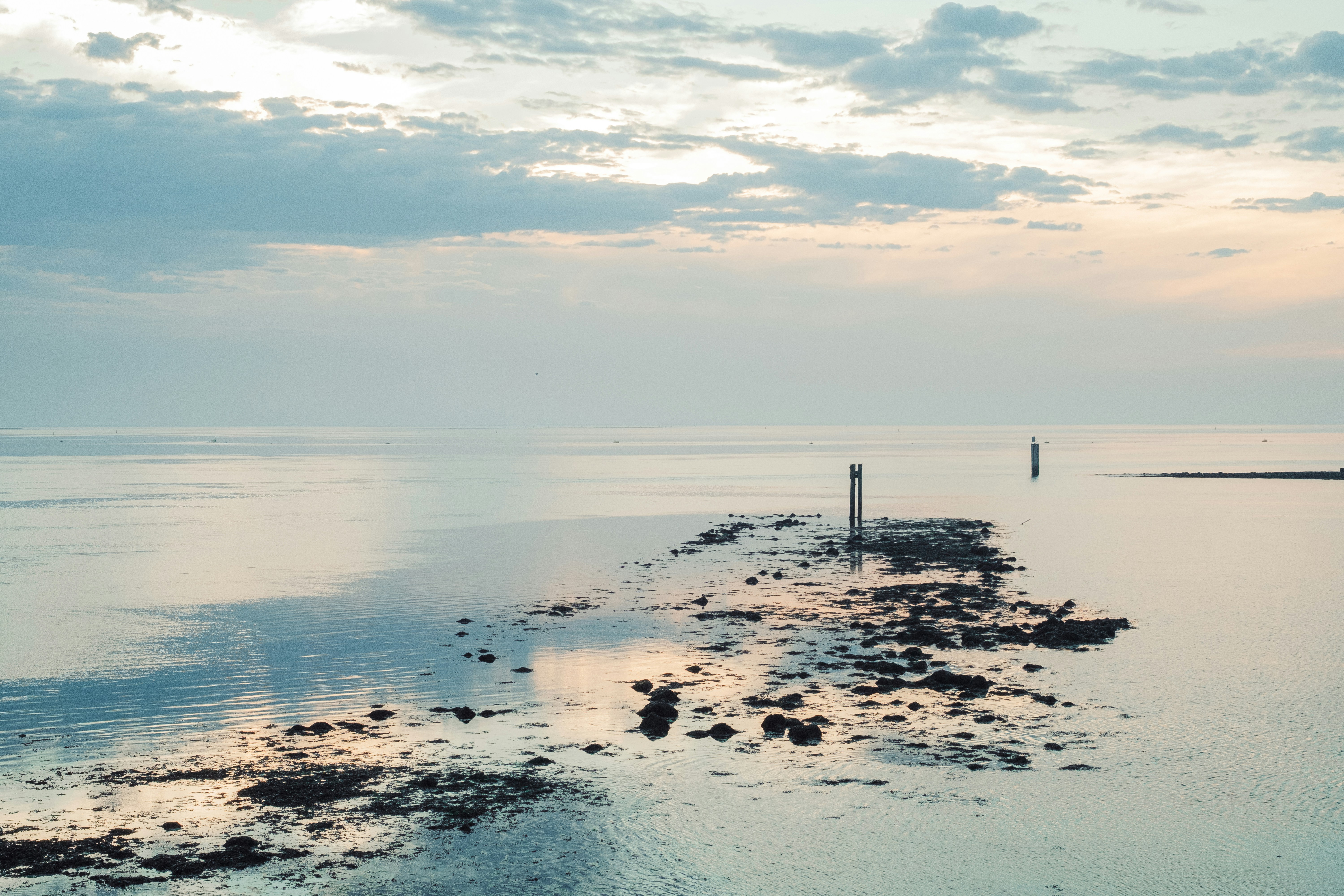 A serene, pastel-toned seascape capturing low tide along the Zeeland coast. An exposed rocky ridge with wooden posts leads out into calm, glassy water that softly reflects a subtle, cloudy sunset sky.