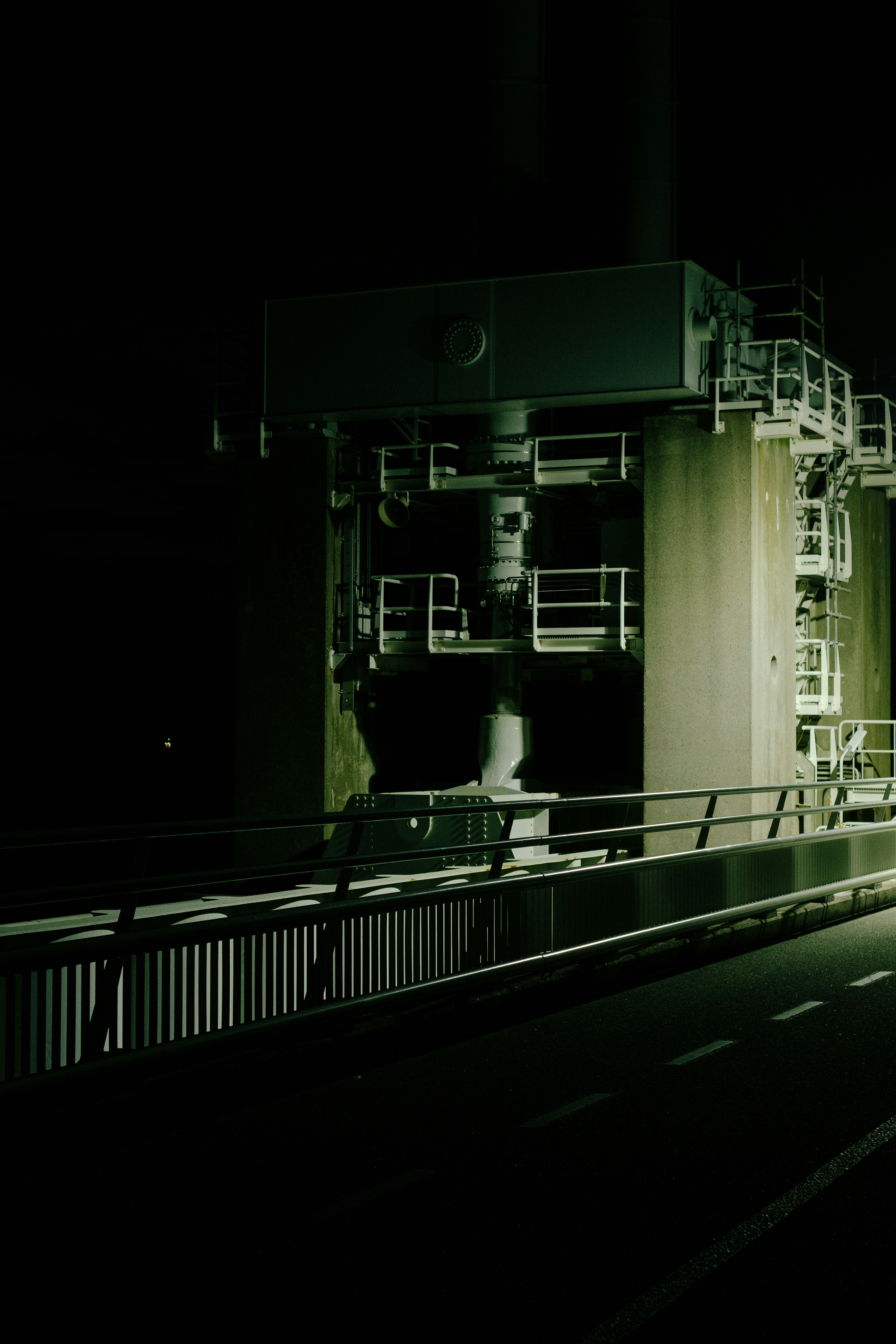 A moody, low-key night photograph of a massive concrete pier and hydraulic gate mechanism along the Oosterscheldekering storm surge barrier. Harsh industrial lighting casts eerie green highlights across the steel railings and brutalist concrete structure against total darkness.