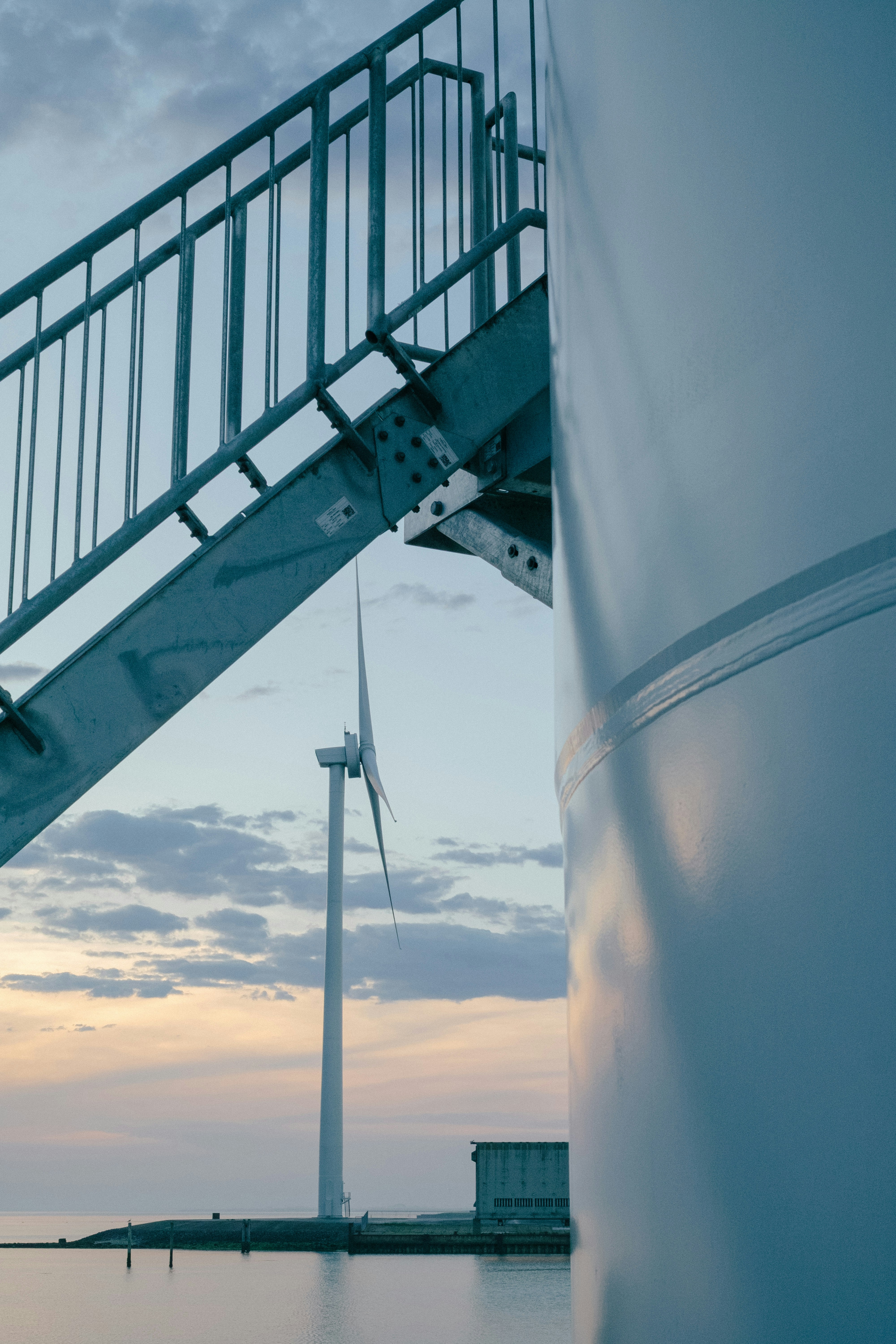 An inventive architectural perspective using the sleek tower and metallic staircase of one wind turbine to frame another standing across the harbor. Set along the Zeeland coast during twilight, soft pastel orange and blue tones fill the evening sky above calm waters.