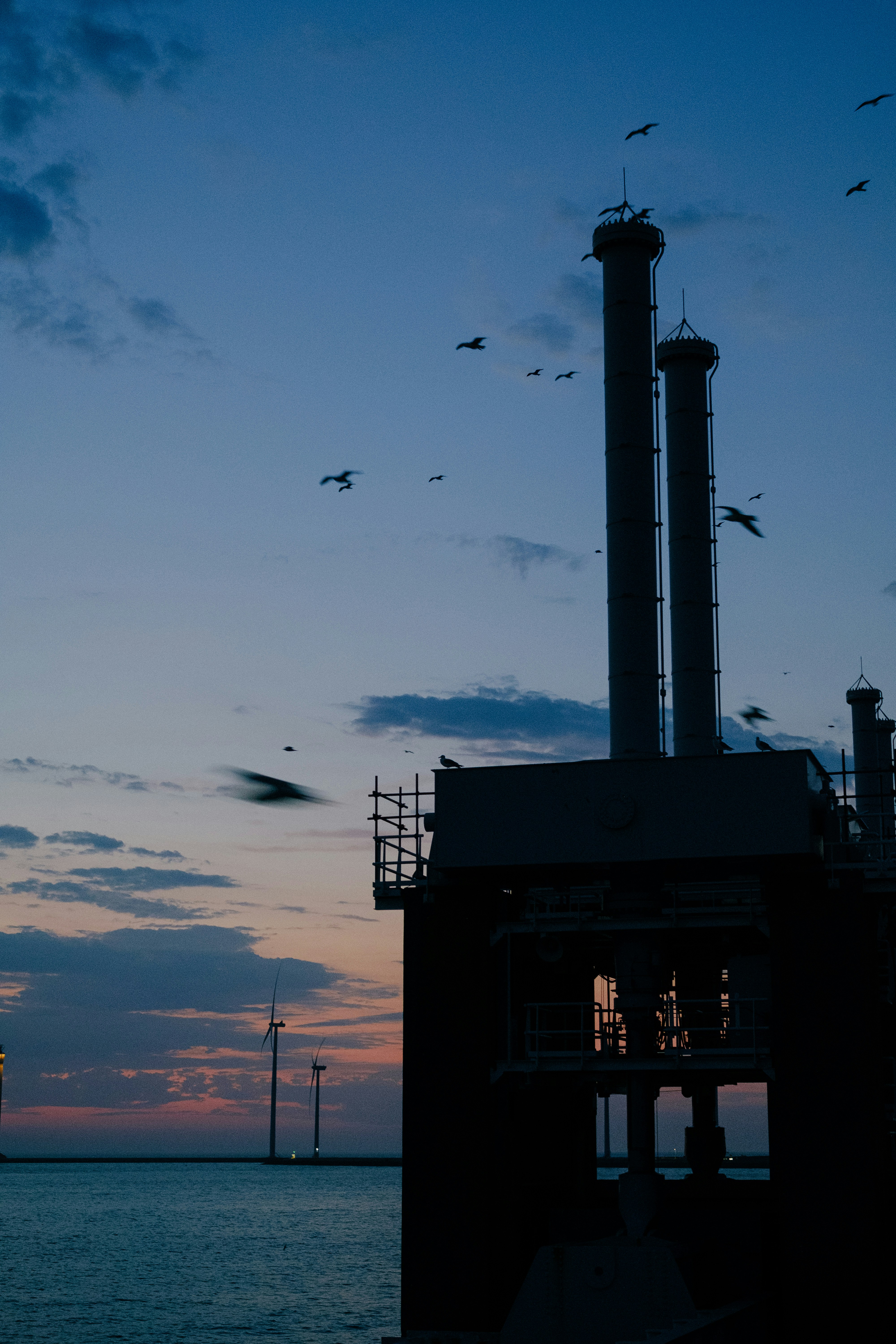 A striking twilight silhouette of the Oosterscheldekering storm surge barrier towering against a deep blue and dusk-orange sky. A flock of seagulls glides around the massive industrial structure, with distant wind turbines standing along the horizon over calm coastal waters.