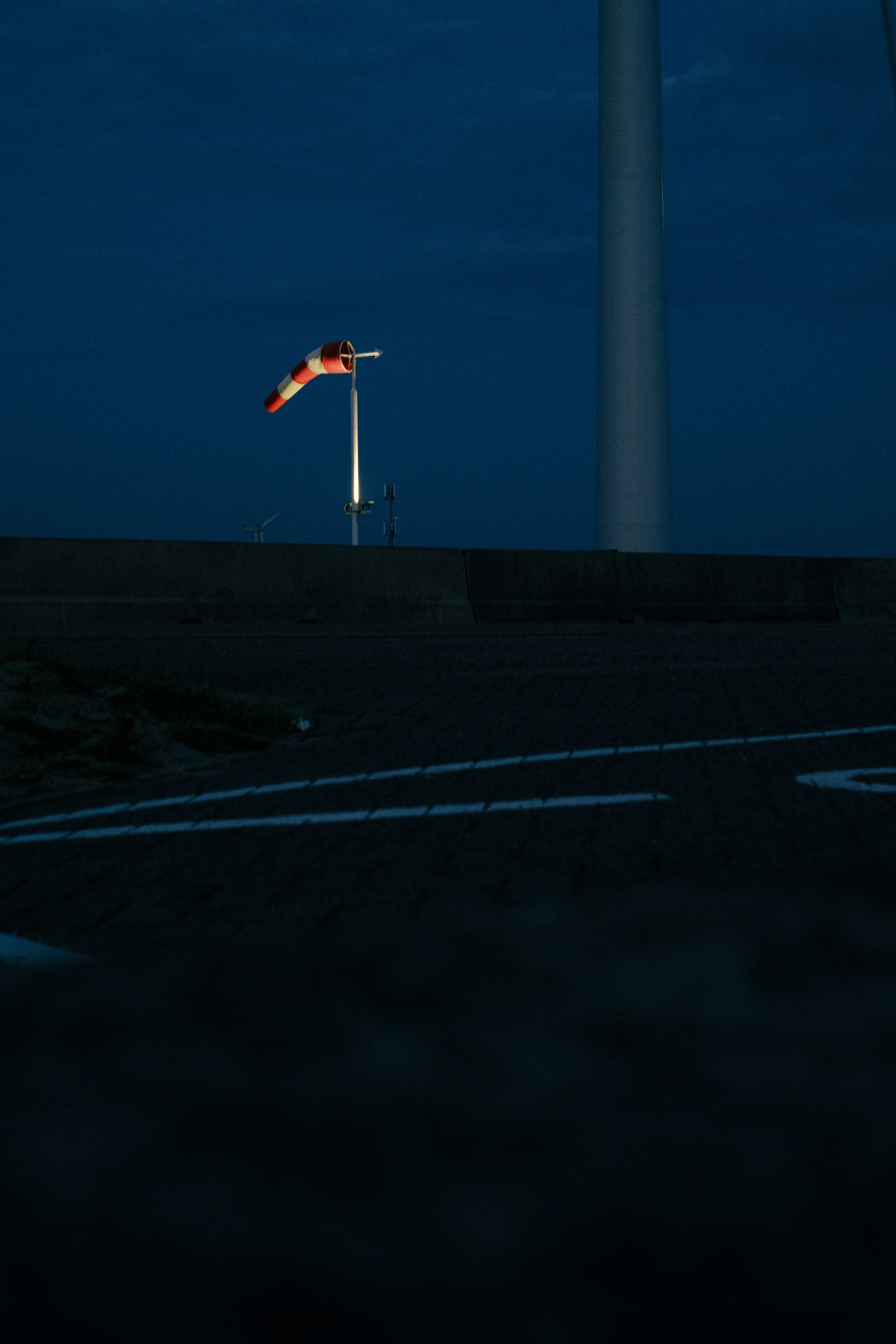 A moody, low-key night photograph of an illuminated red-and-white windsock standing along the Zeeland coast. Set against a deep blue hour sky and the shadowy silhouette of a towering wind turbine, the upward spotlight creates a vivid, minimalist focal point in the dark landscape.