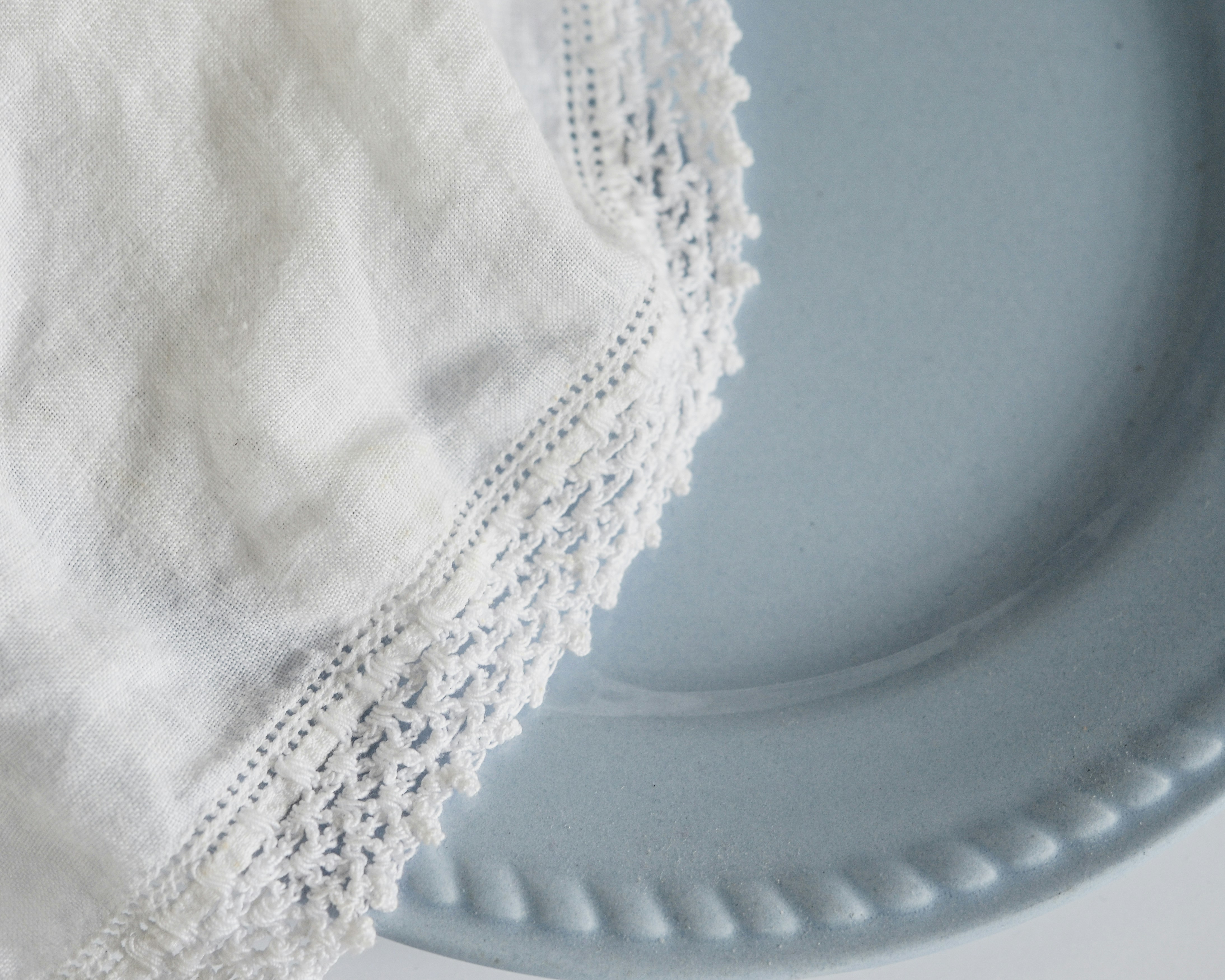 Close-up of a delicate lace edge resting on the rim of a pale blue plate, emphasizing texture and contrast between fabric and ceramic. (Logo/plate)