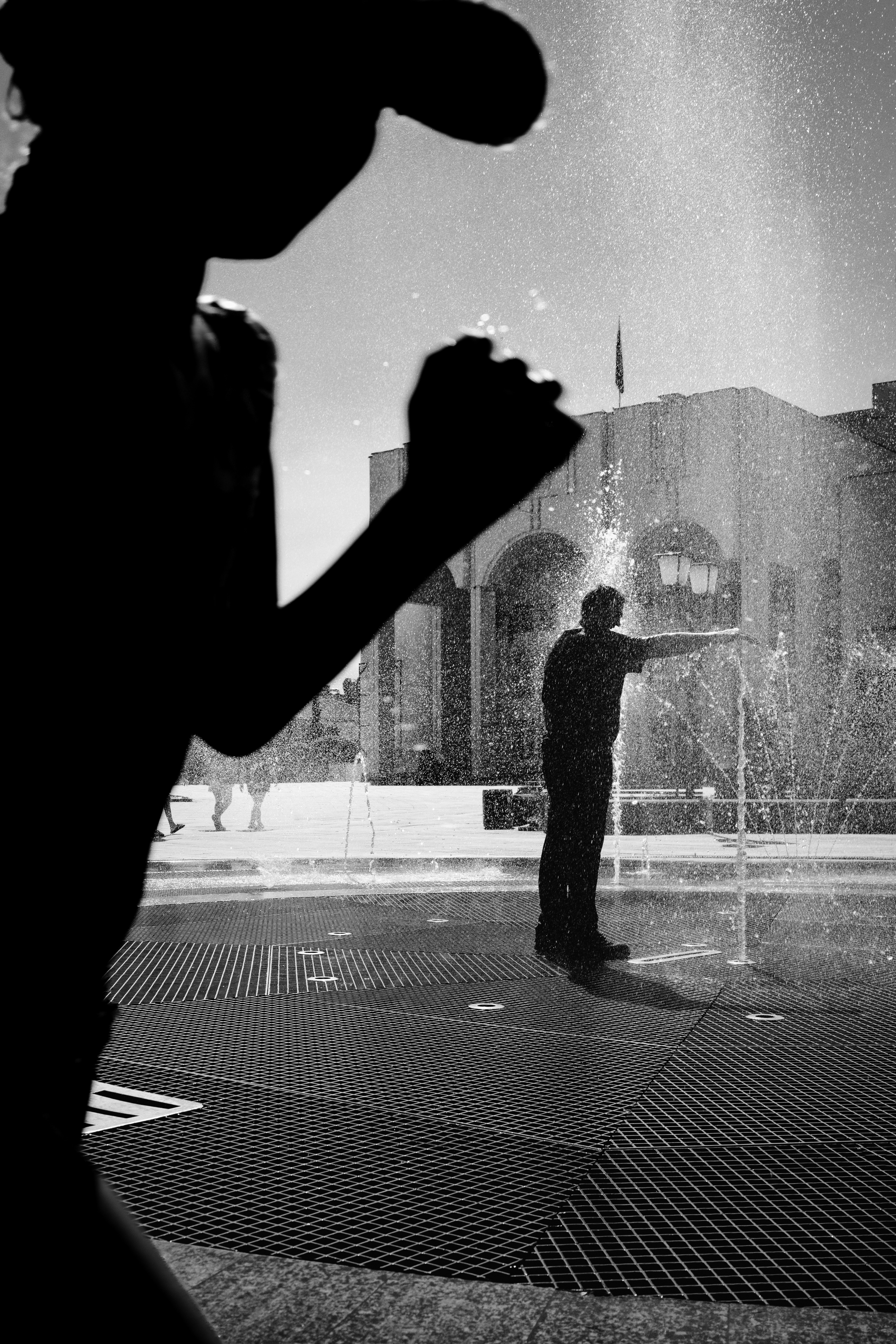 High-contrast black-and-white scene of a silhouetted figure in the foreground with a water fountain and distant architected plaza; a secondary figure gestures near a spray jet, hinting at motion and interaction.