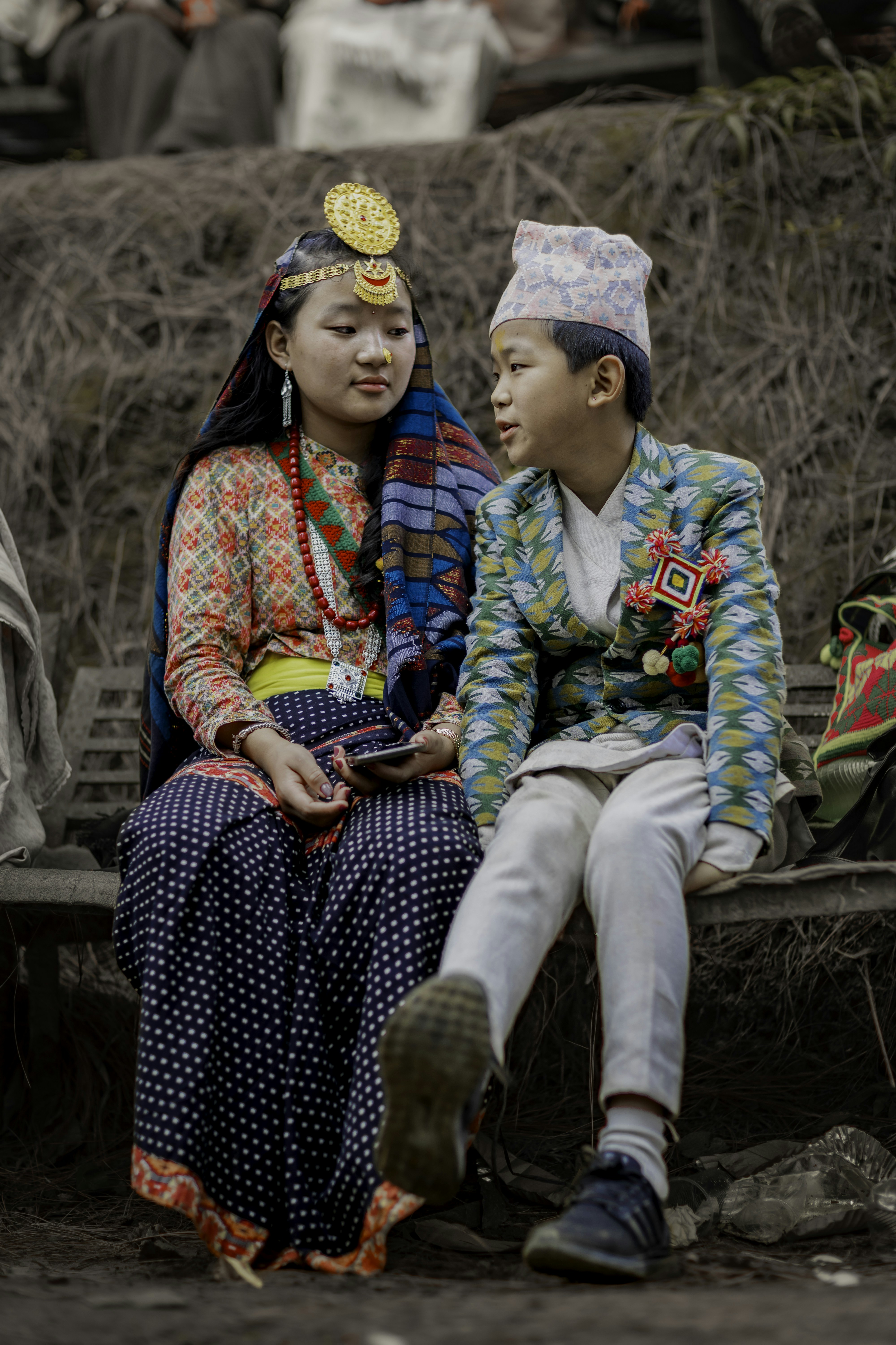Two young performers in colorful traditional attire share a quiet moment on a rustic bench