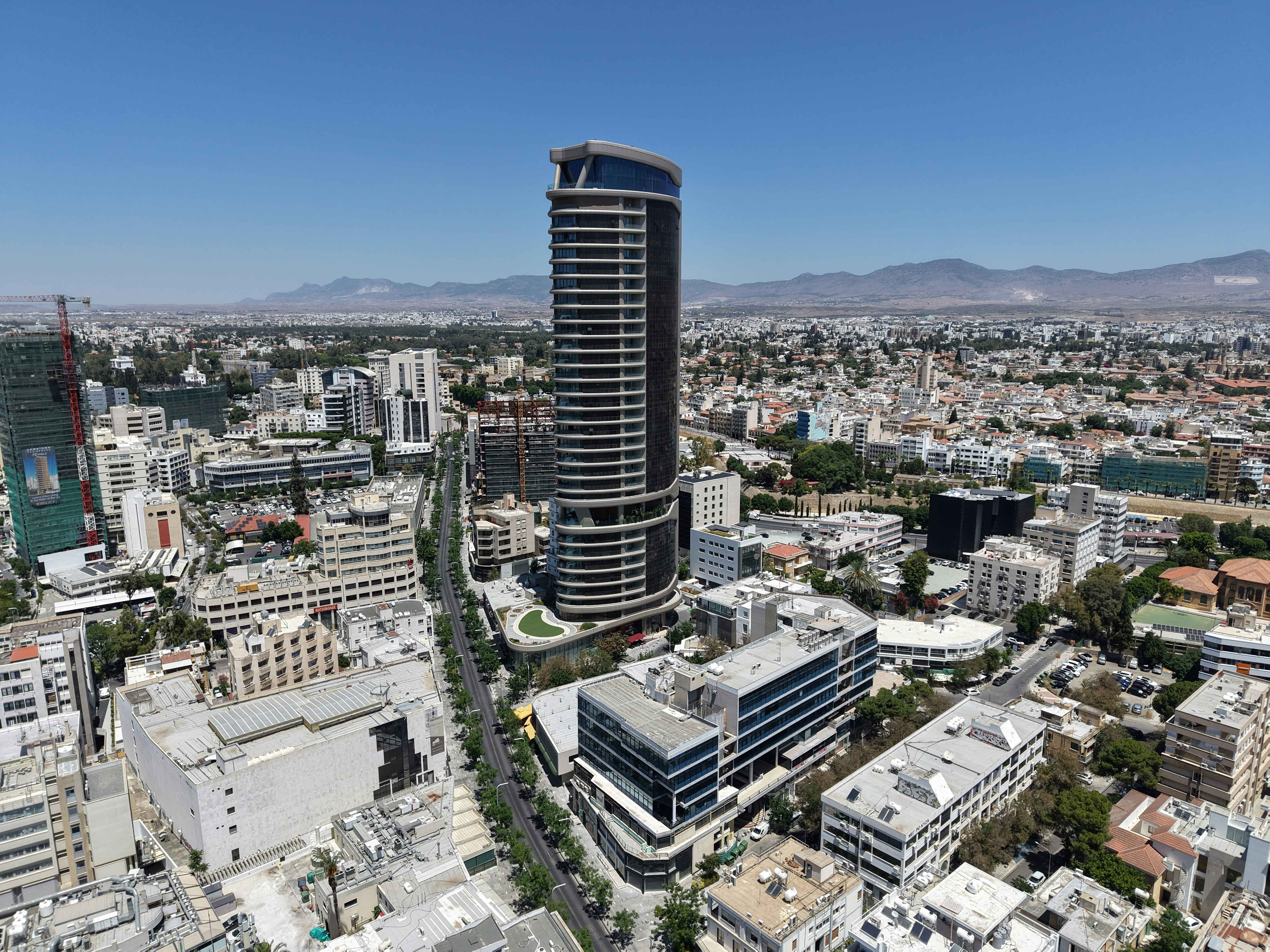 Aerial view of a dense urban core with a prominent modern skyscraper rising among lower buildings; the scene emphasizes a geometric city grid under a clear blue sky.
