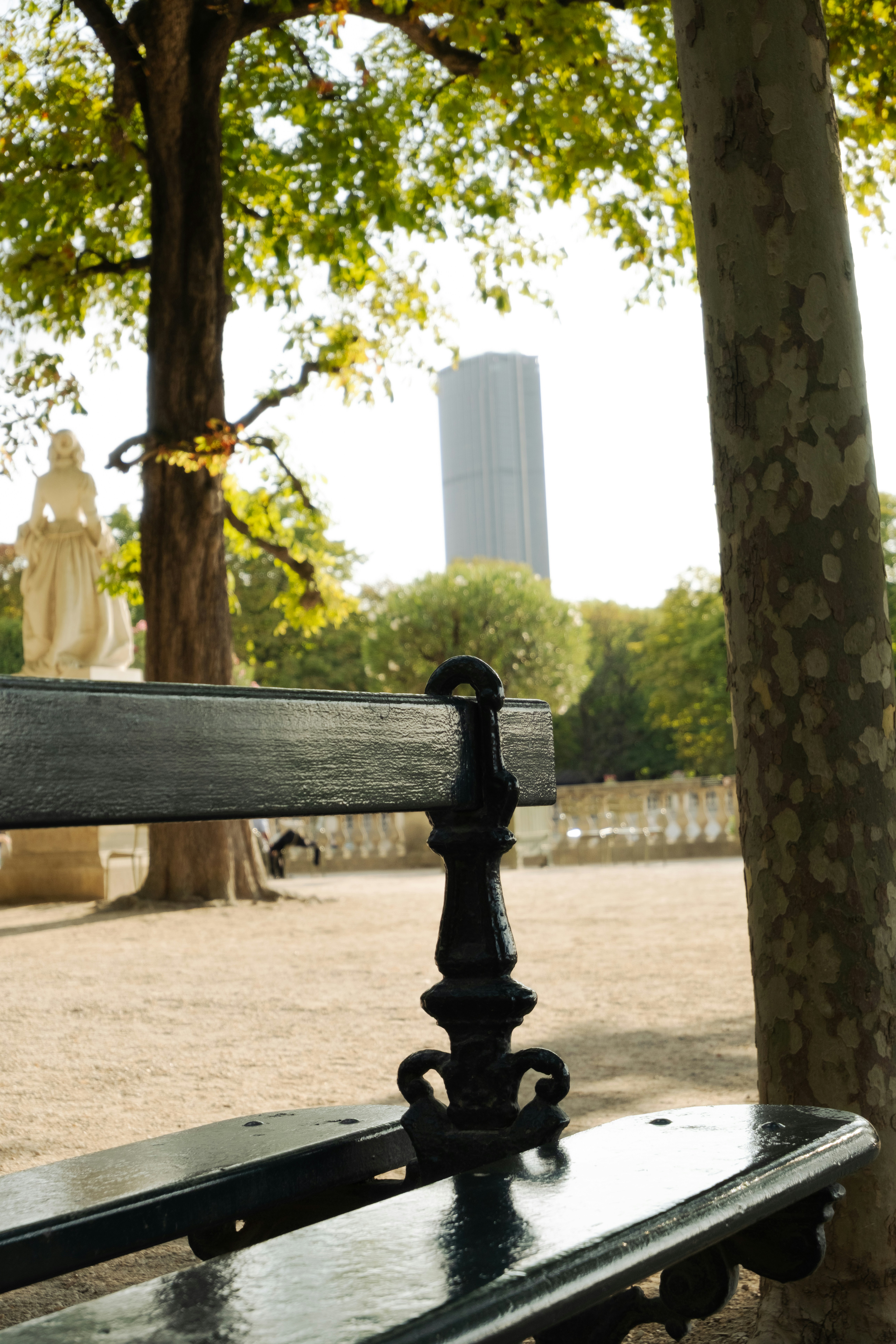 Sunlit park bench with ornate ironwork in the foreground; a statue and distant skyscraper rise beyond trees in the background, suggesting a tranquil urban garden scene.