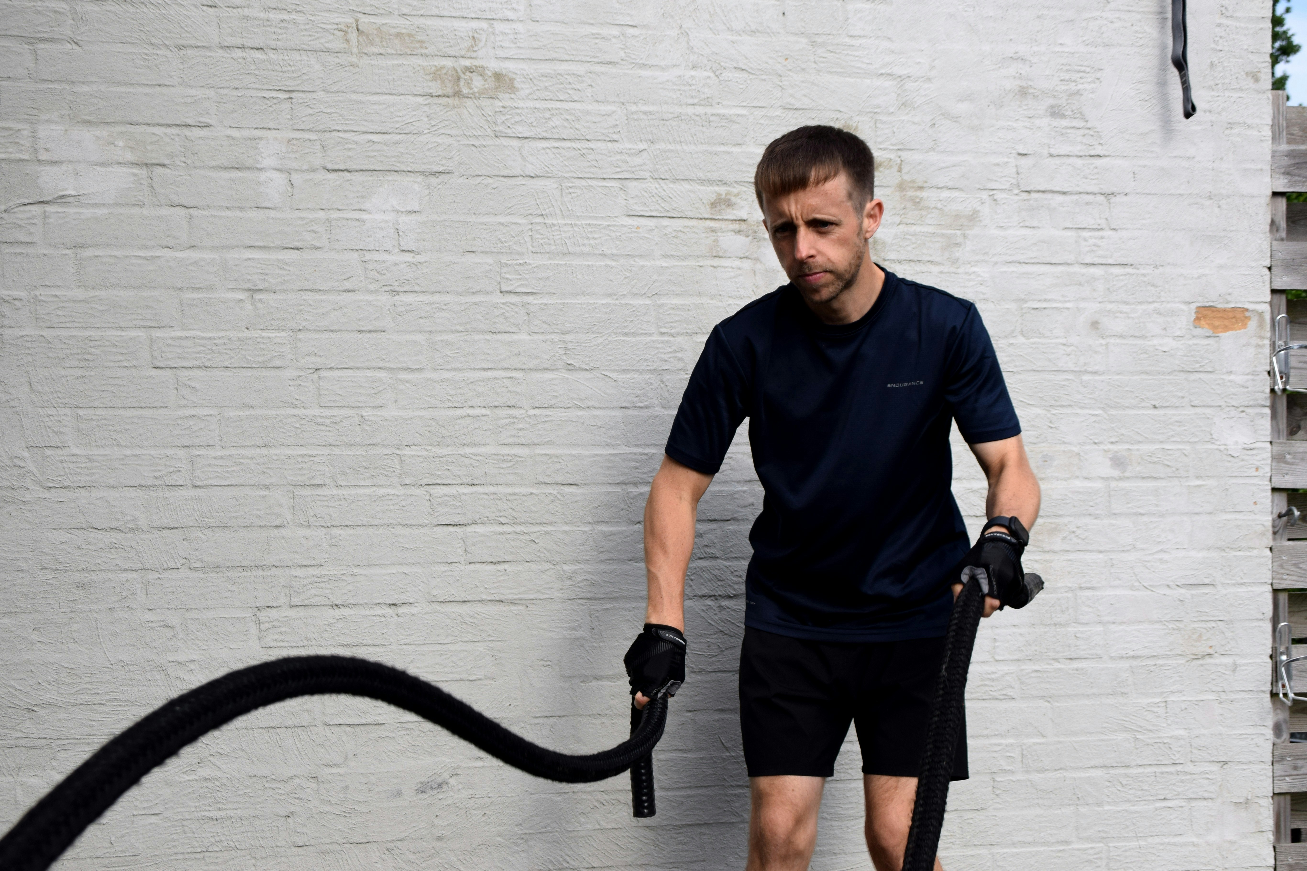 Rope Workout: Man in navy apparel grips a thick hose against a pale brick wall