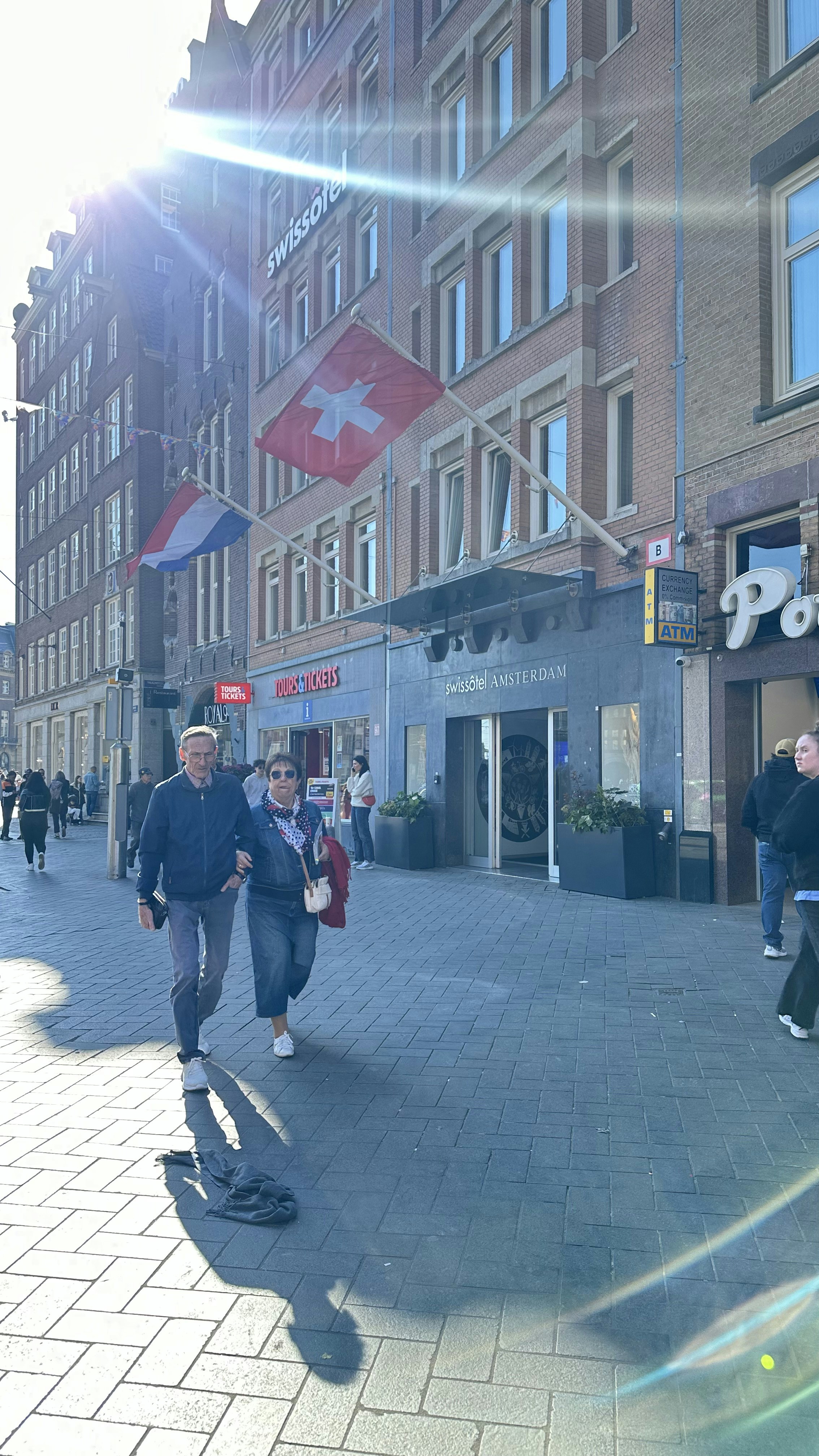 Bustling city street with Swiss and Dutch flags above a row of storefronts, pedestrians strolling on a sunlit, tiled pavement.