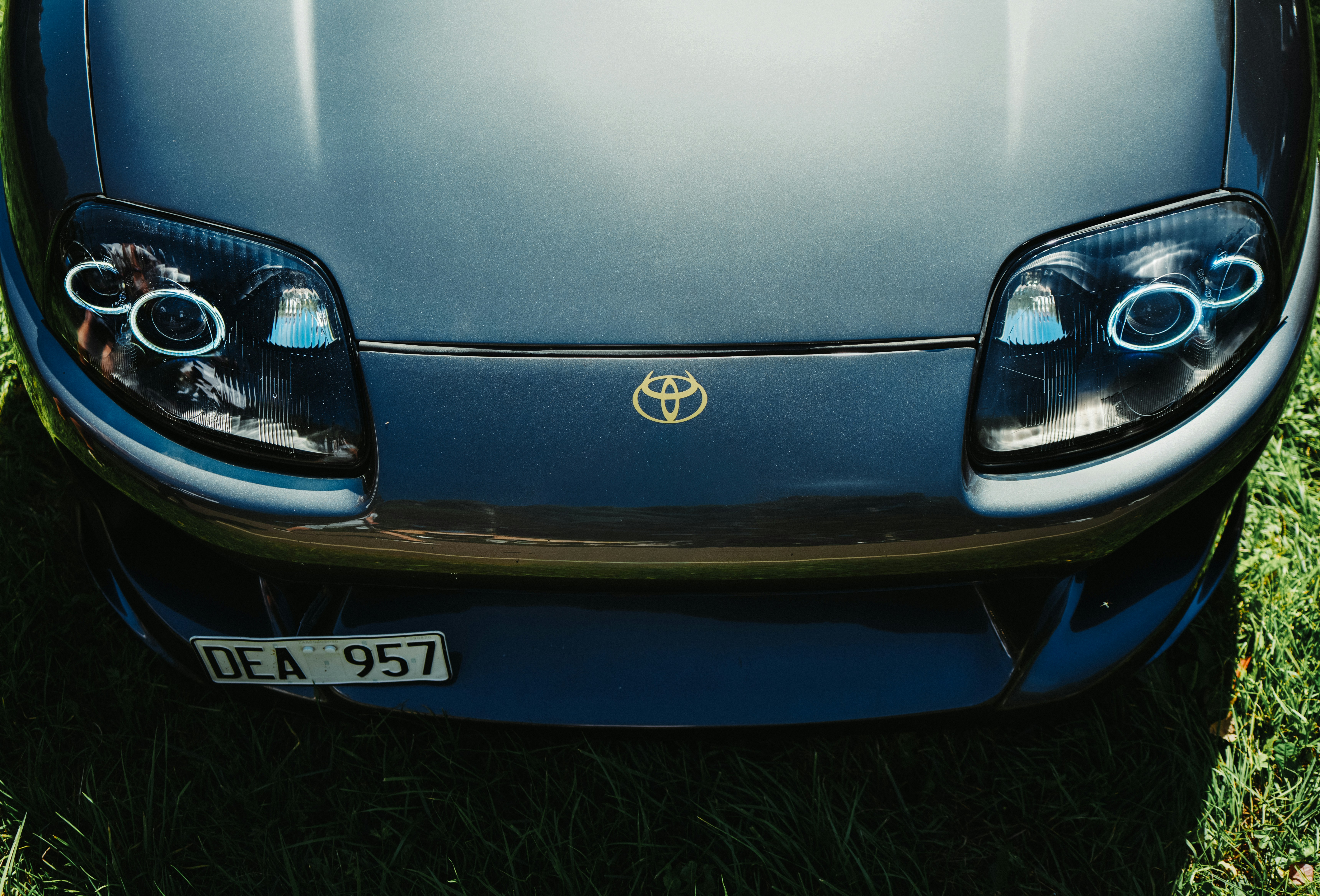 Front-view of a blue Toyota-labeled sports coupe with sunlit headlights and a grassy foreground
