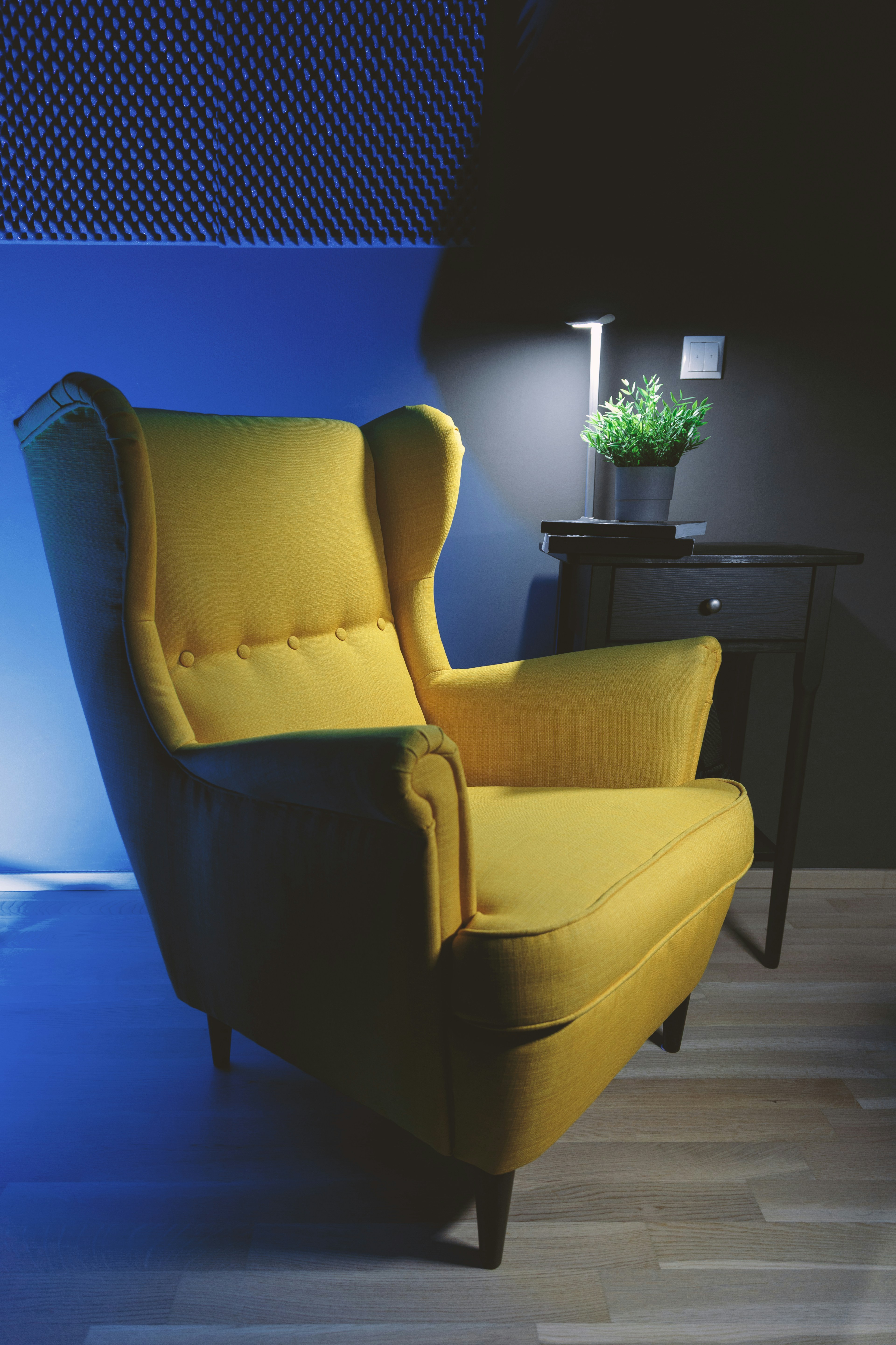 A vibrant yellow armchair sits in a moody, modern studio corner with a small side table and plant; soft lighting highlights the chair against a dark wall and blue backdrop. The scene resembles a staged interior setup.