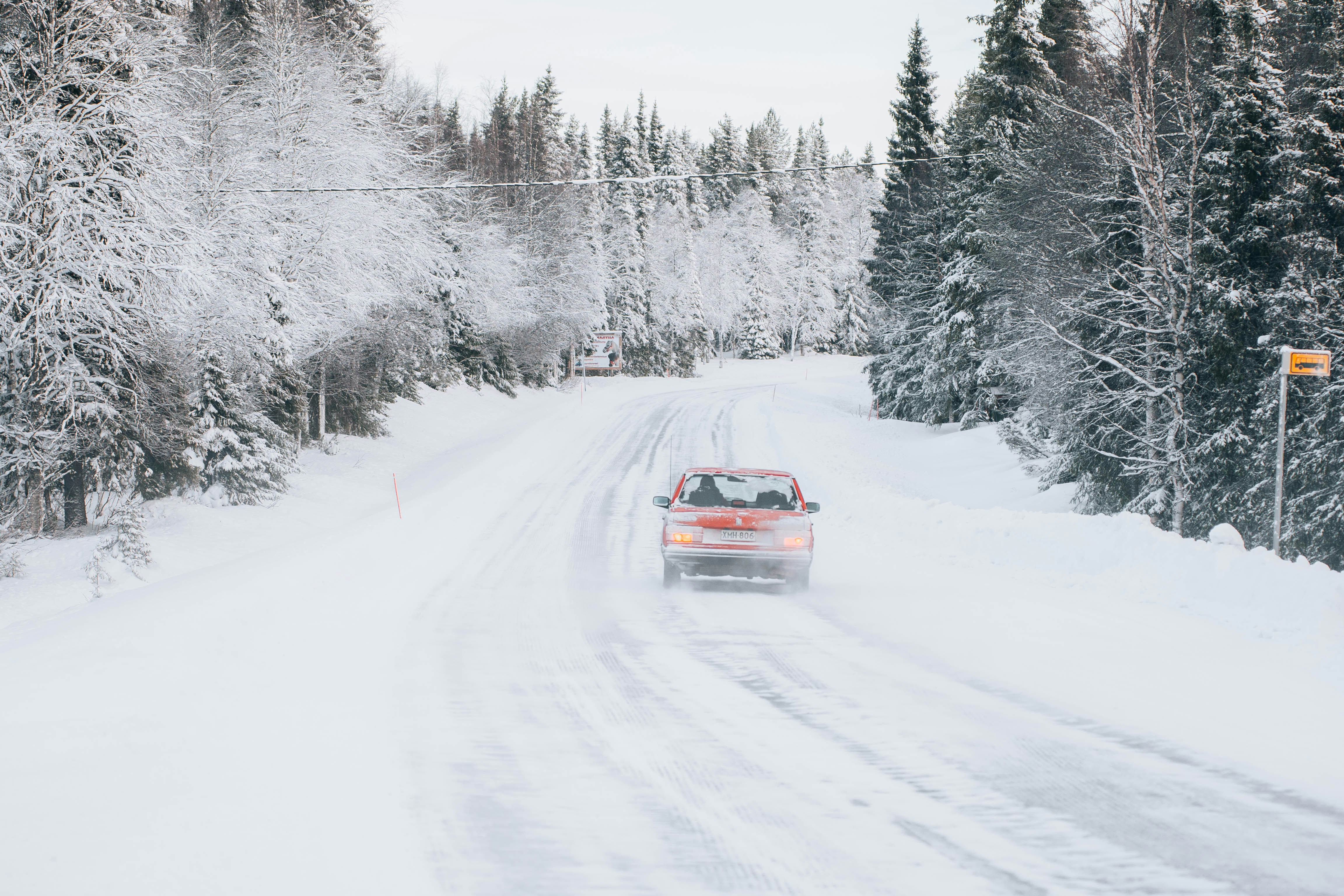 A lone red car drives down a snow-covered rural road flanked by frosted trees, embodying a quiet winter journey. Snow blankets the scene as tire tracks mark the path ahead.