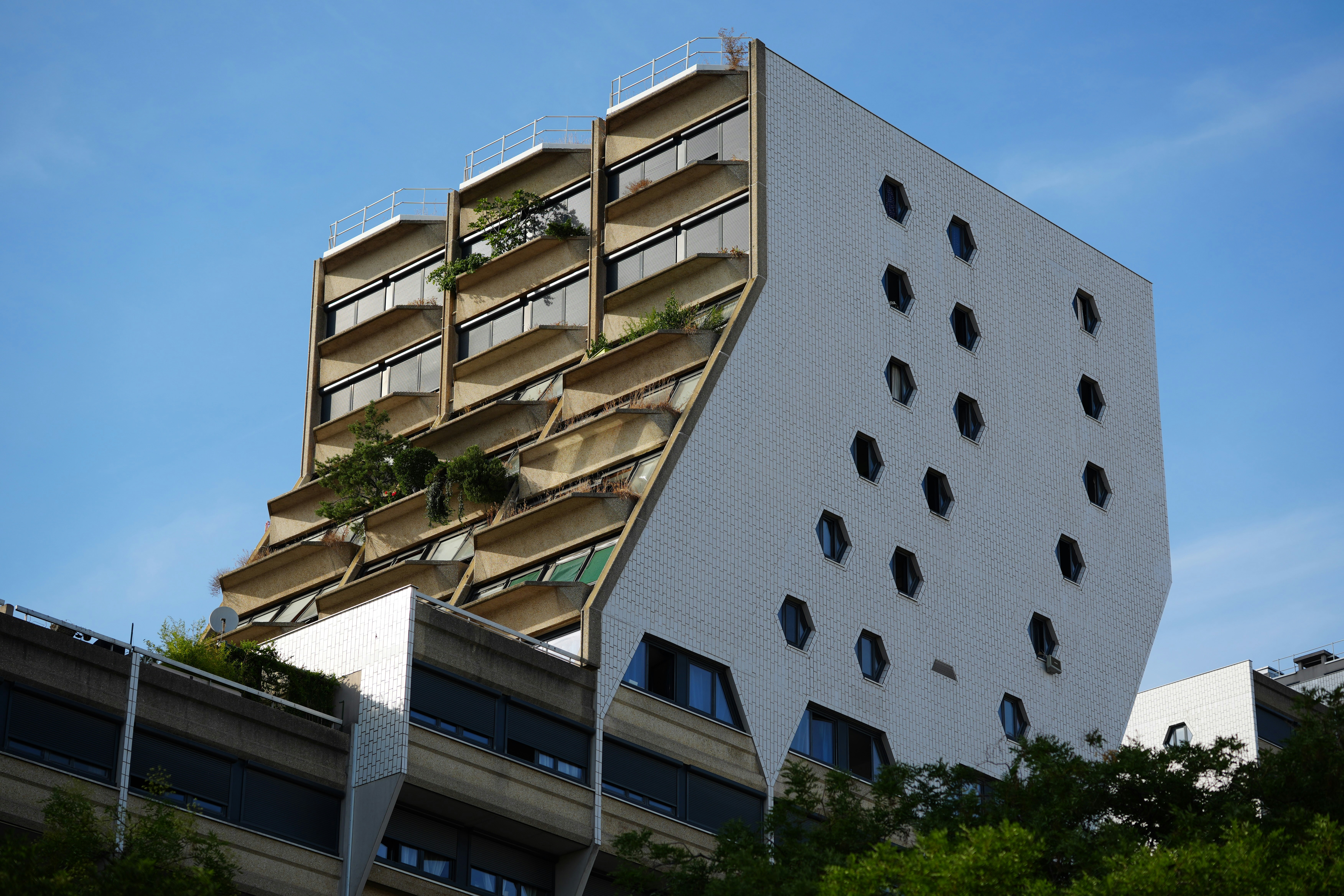 Architectural composition featuring a modern, angular building with a stepped, perforated facade and a slanted white wall dotted with octagonal windows; foreground shows lower levels and greenery. A study in geometric repetition and urban texture.