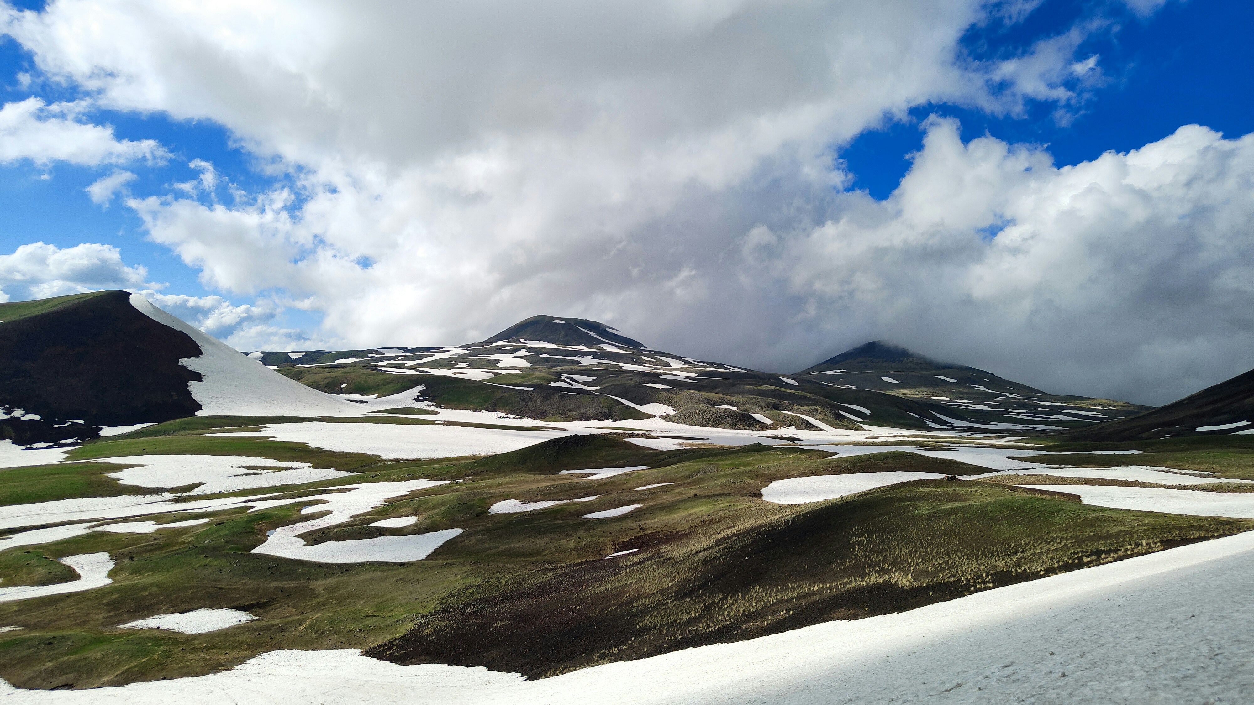 Snow patches weave through mossy hillside under dramatic clouds, a remote alpine landscape with distant peaks and a vivid blue sky.
