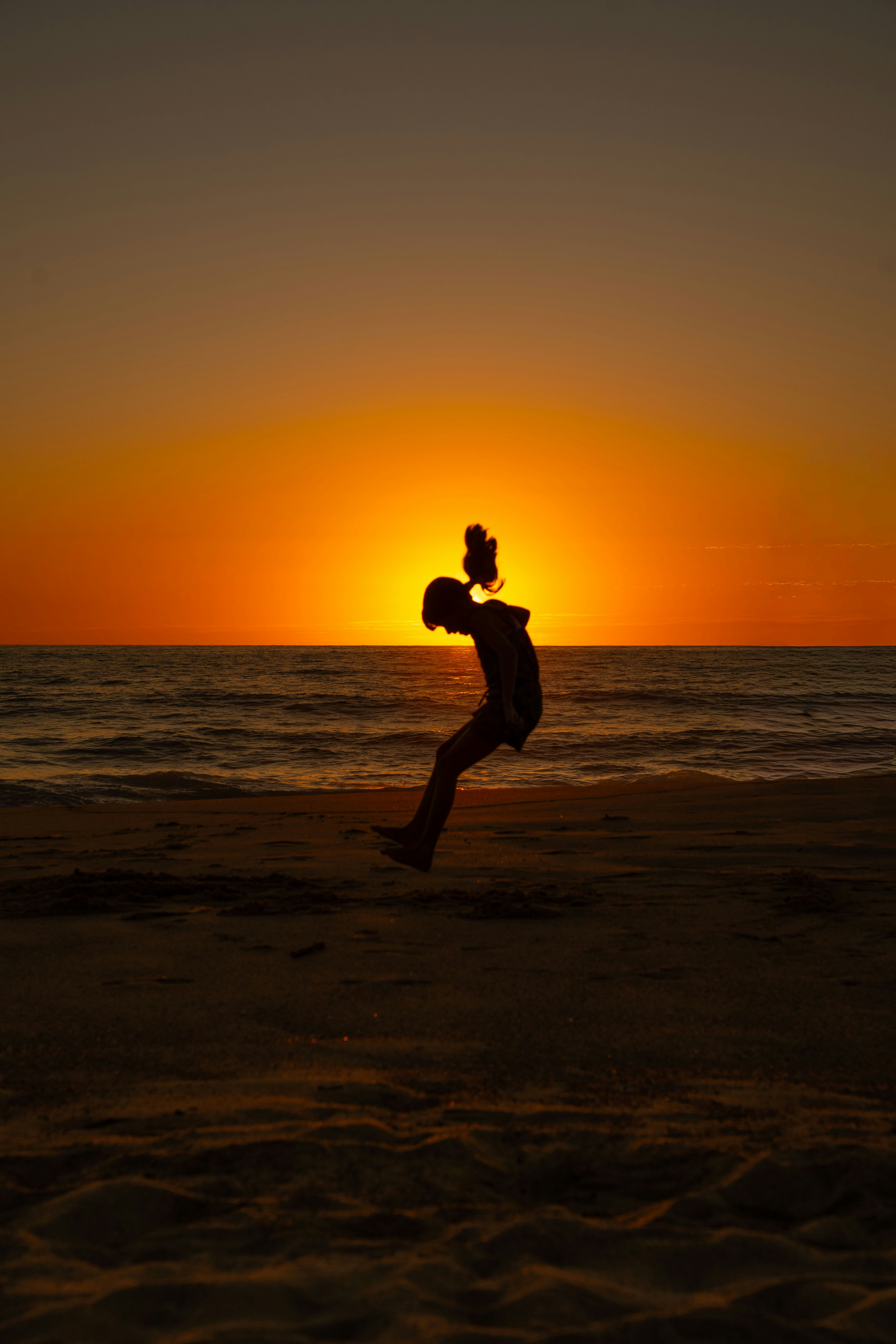 Silhouette of a person mid-jump on a beach during sunset, with the sun low on the horizon and warm orange tones across the sky.