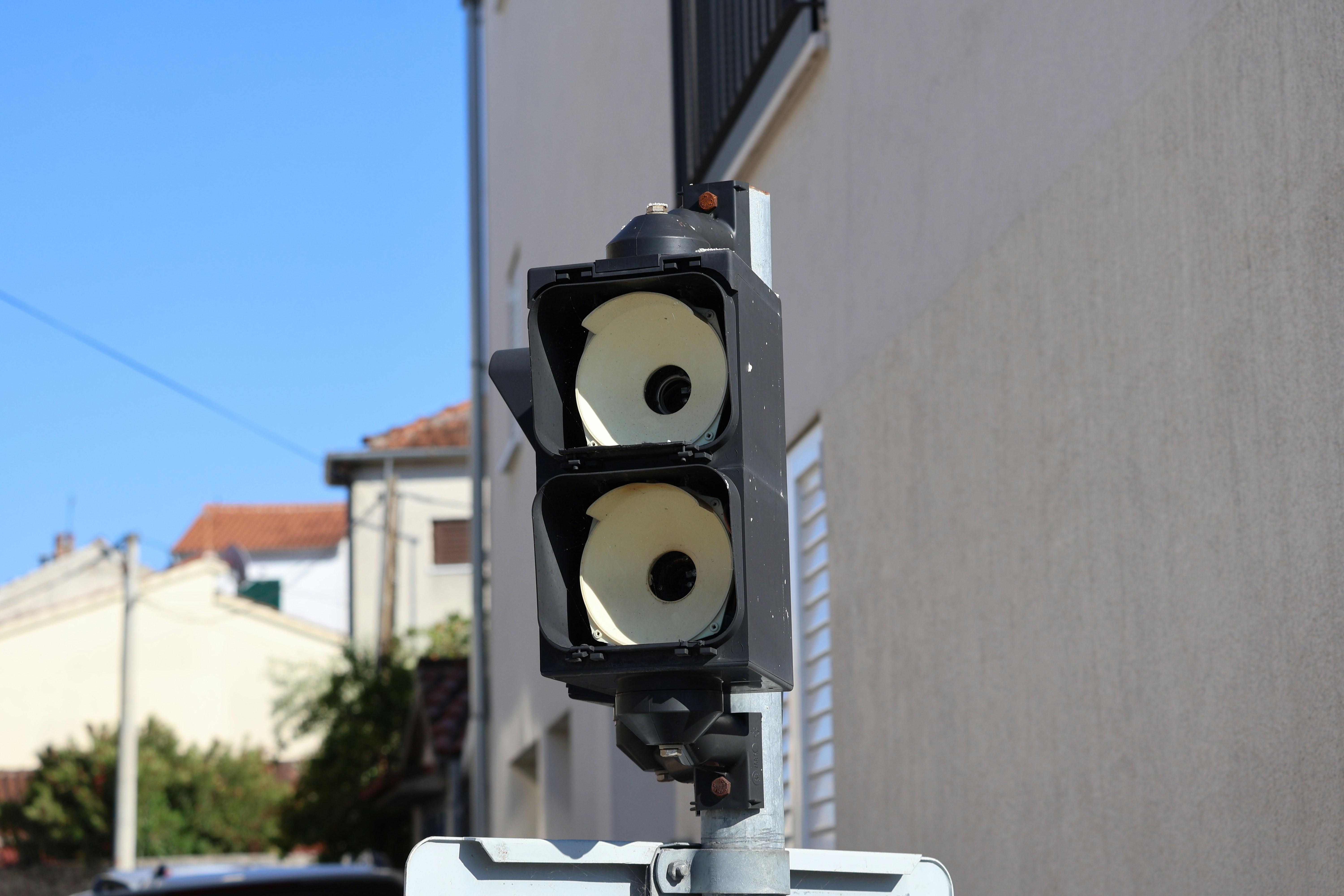 A close-up of a broken traffic light with missing lenses, revealing the empty reflective cones inside. The object stands in sharp focus against a clear blue sky and blurred European-style buildings, conveying a mood of urban neglect on a bright sunny day