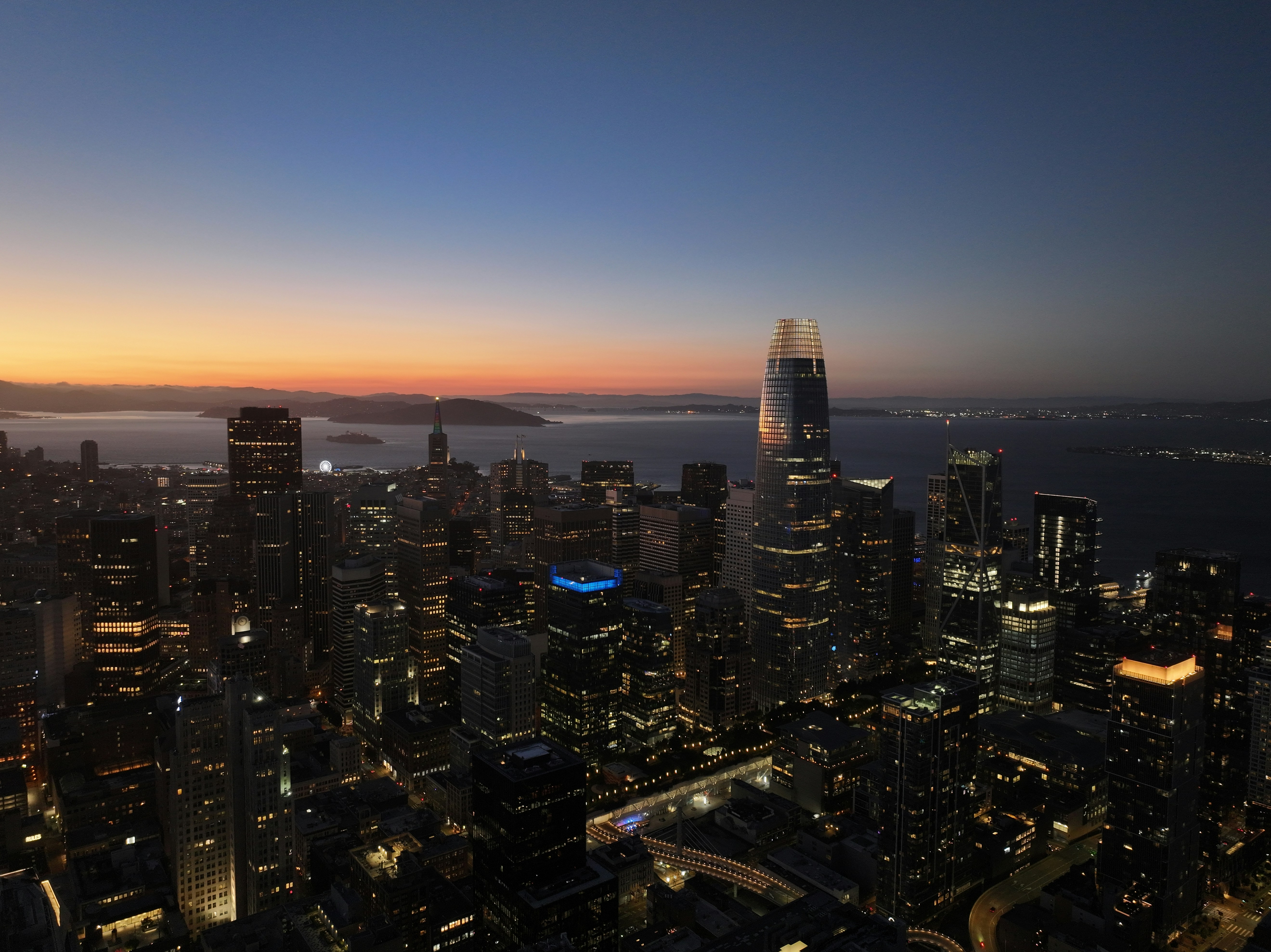 A nocturnal cityscape perched along a coastline, with clustered high-r rises glowing against a fading sunset and a lone tall tower standing out among the skyline.