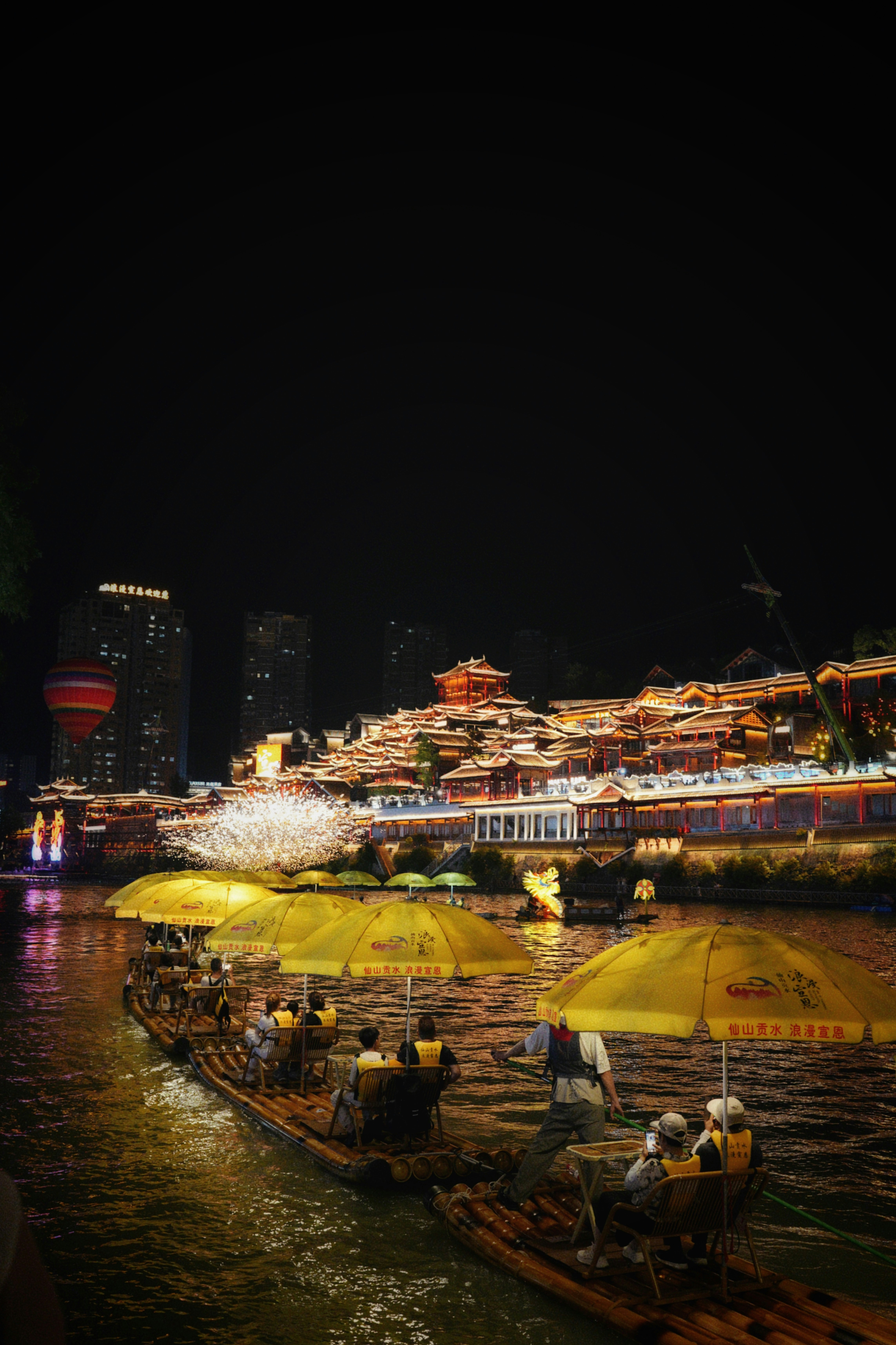 Night scene of a river boat with yellow umbrellas gliding under a brightly lit riverside with traditional architecture; a cityscape and fireworks visible in the distance.