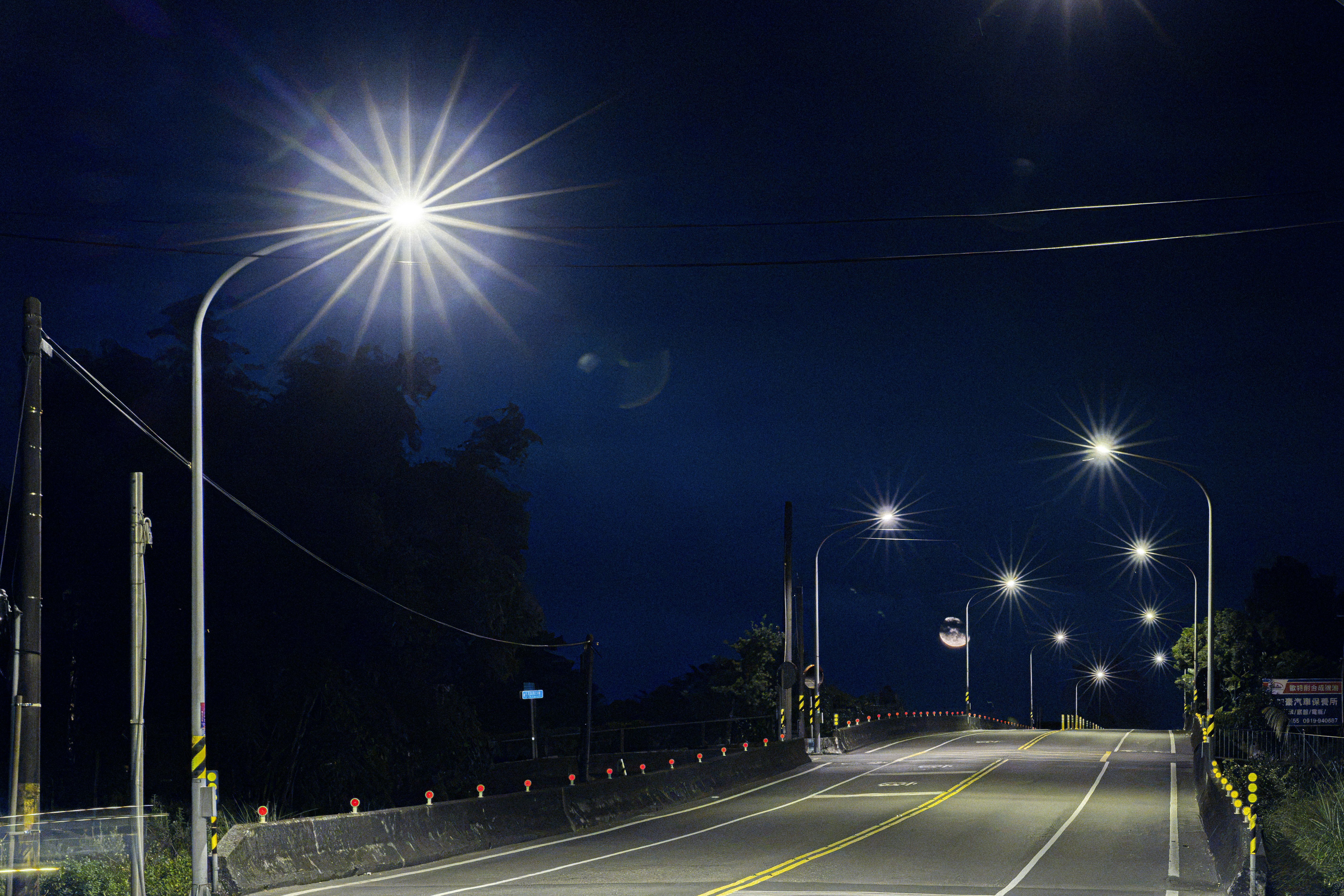 Nighttime street scene with a quiet road lined by bright streetlights and a starburst flare, capturing long-exposure glow and distant headlights.