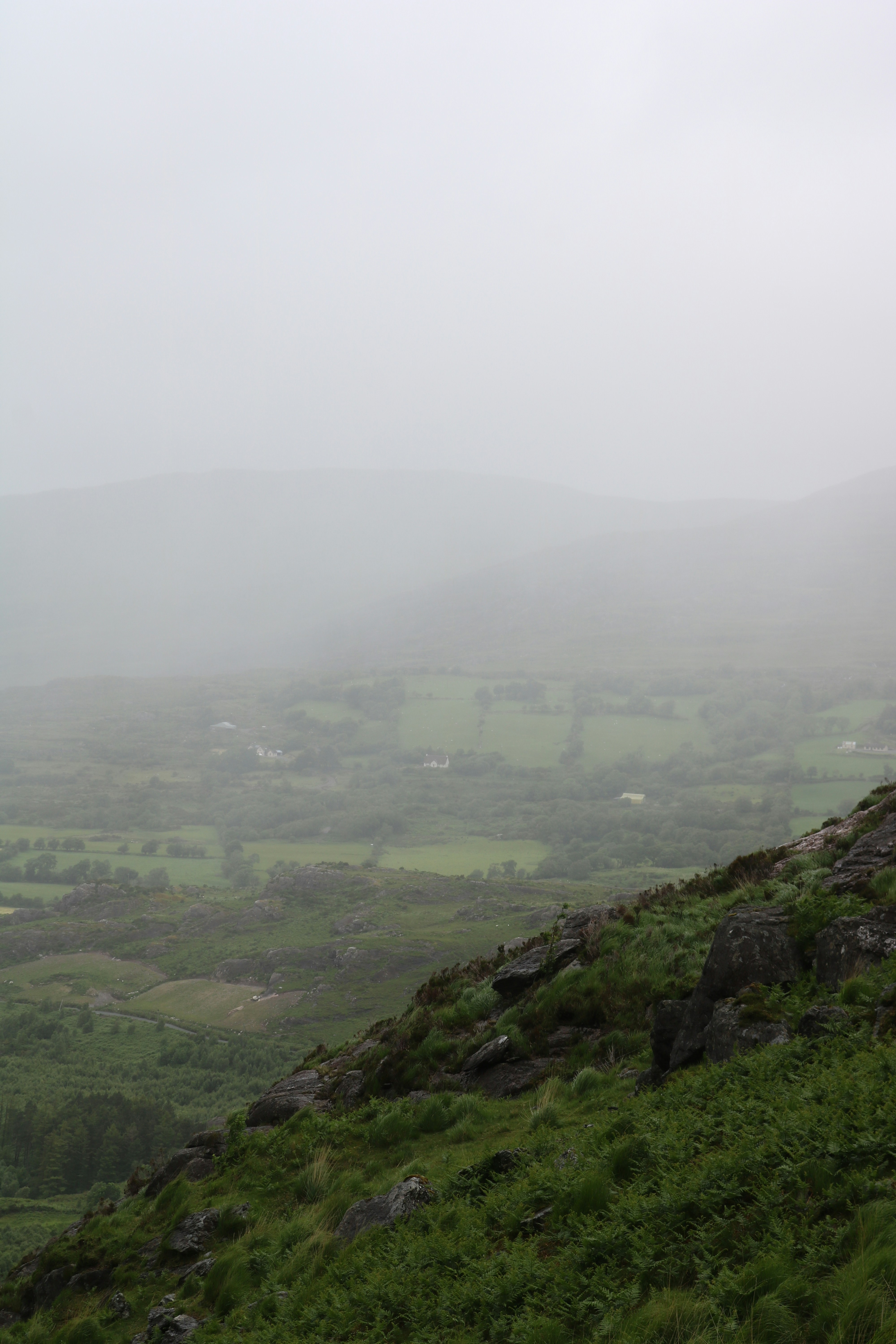 Hazy valley seen from a rocky hillside, with patchwork fields fading into soft fog and distant hills obscured by mist.