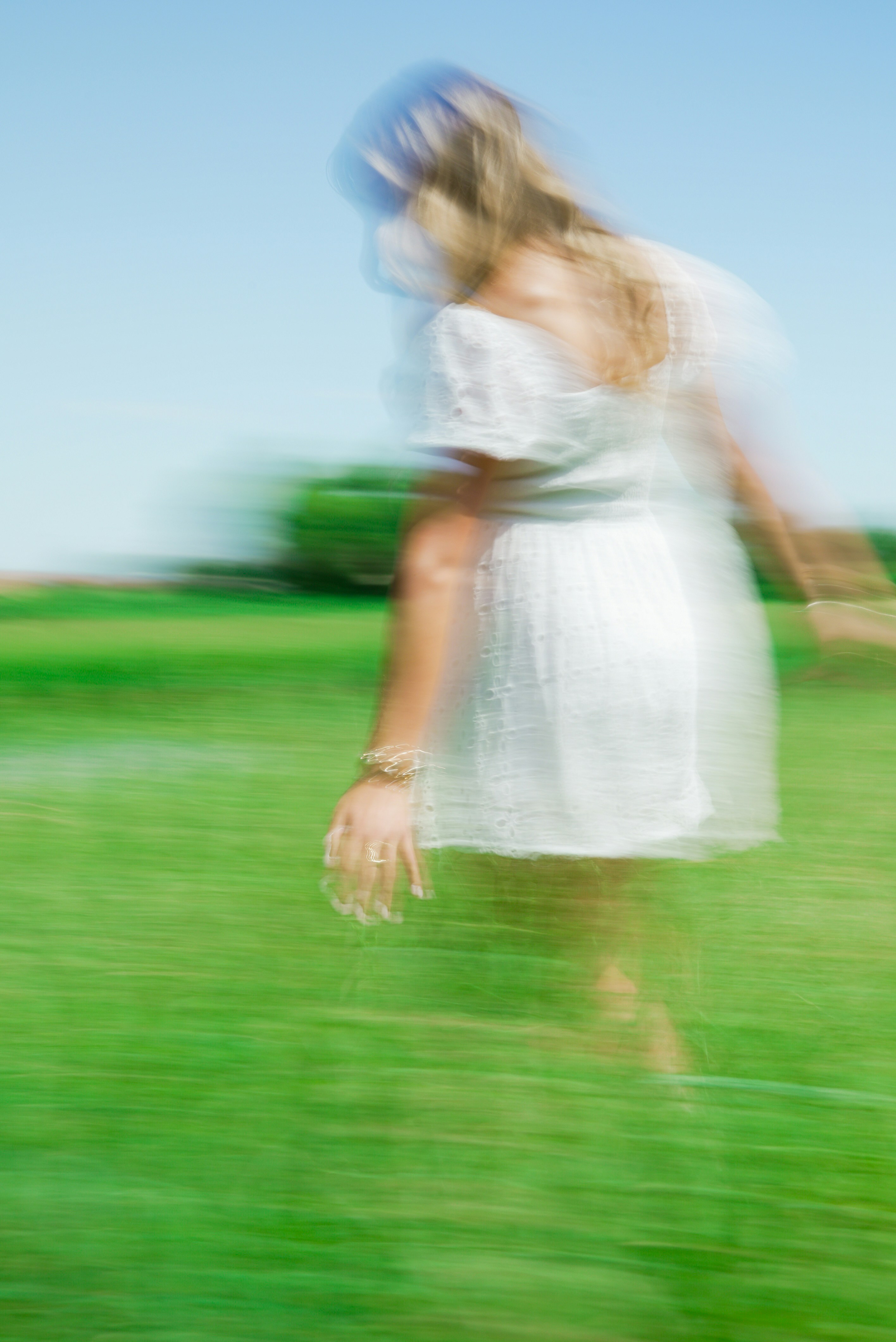 Motion-blurred figure in a white dress moves through a sunlit green field, with a pale blue sky above, creating an abstract, dreamy scene. This is a photograph.