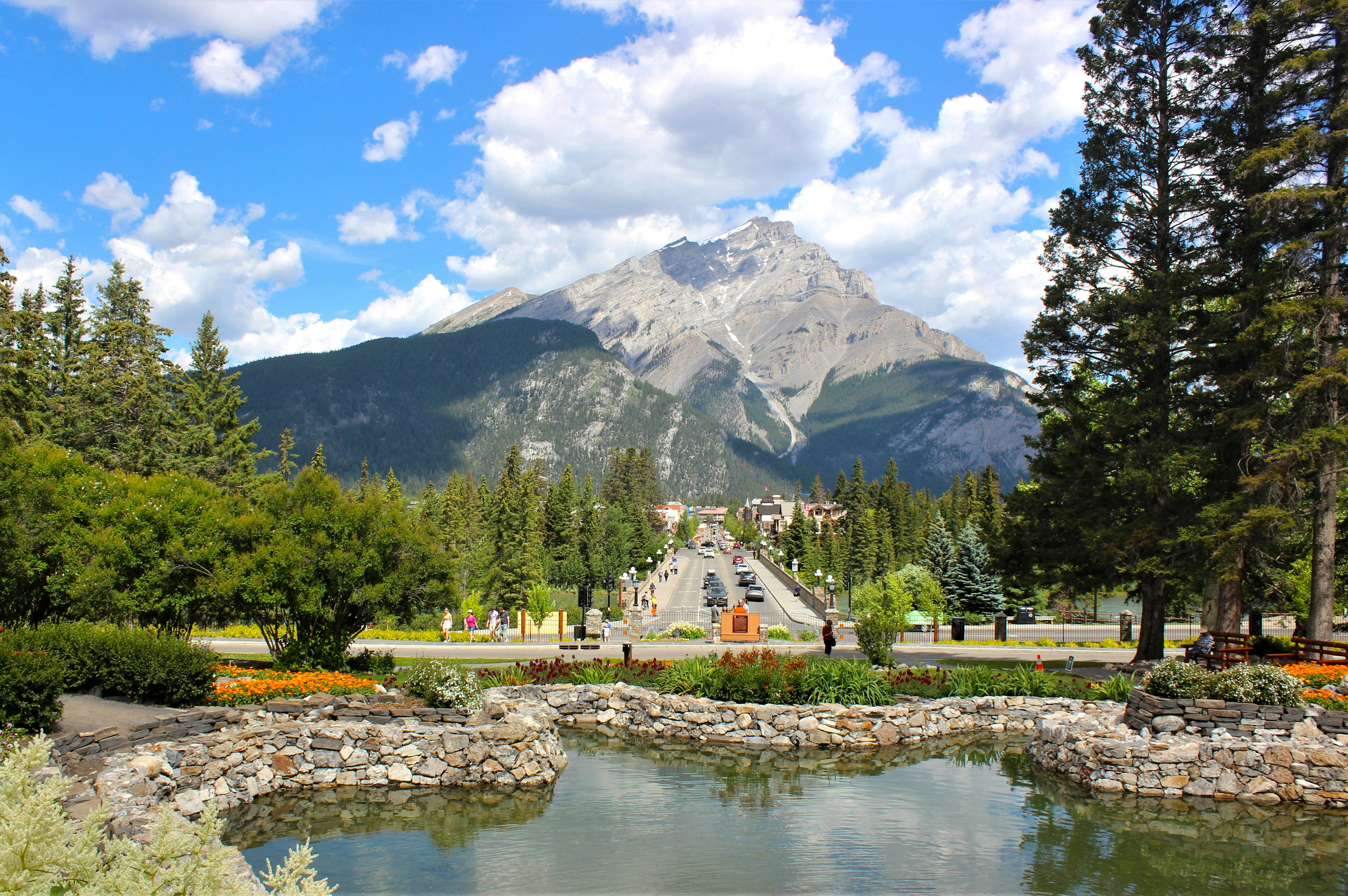 This beautiful landscape captures the iconic view of Cascade Mountain looming directly over the town of Banff, Alberta. The photo is taken from the upper terraces of the Cascade of Time Garden, looking north across a stone-lined reflection pond. Lush green pine trees, vibrant orange and white flower beds, and meticulous stonework frame the bottom of the view. In the middle ground, the Bow River bridge and traffic on Banff Avenue are visible, drawing the eye directly toward the massive, rugged limestone peak under a bright blue sky filled with fluffy white clouds.
