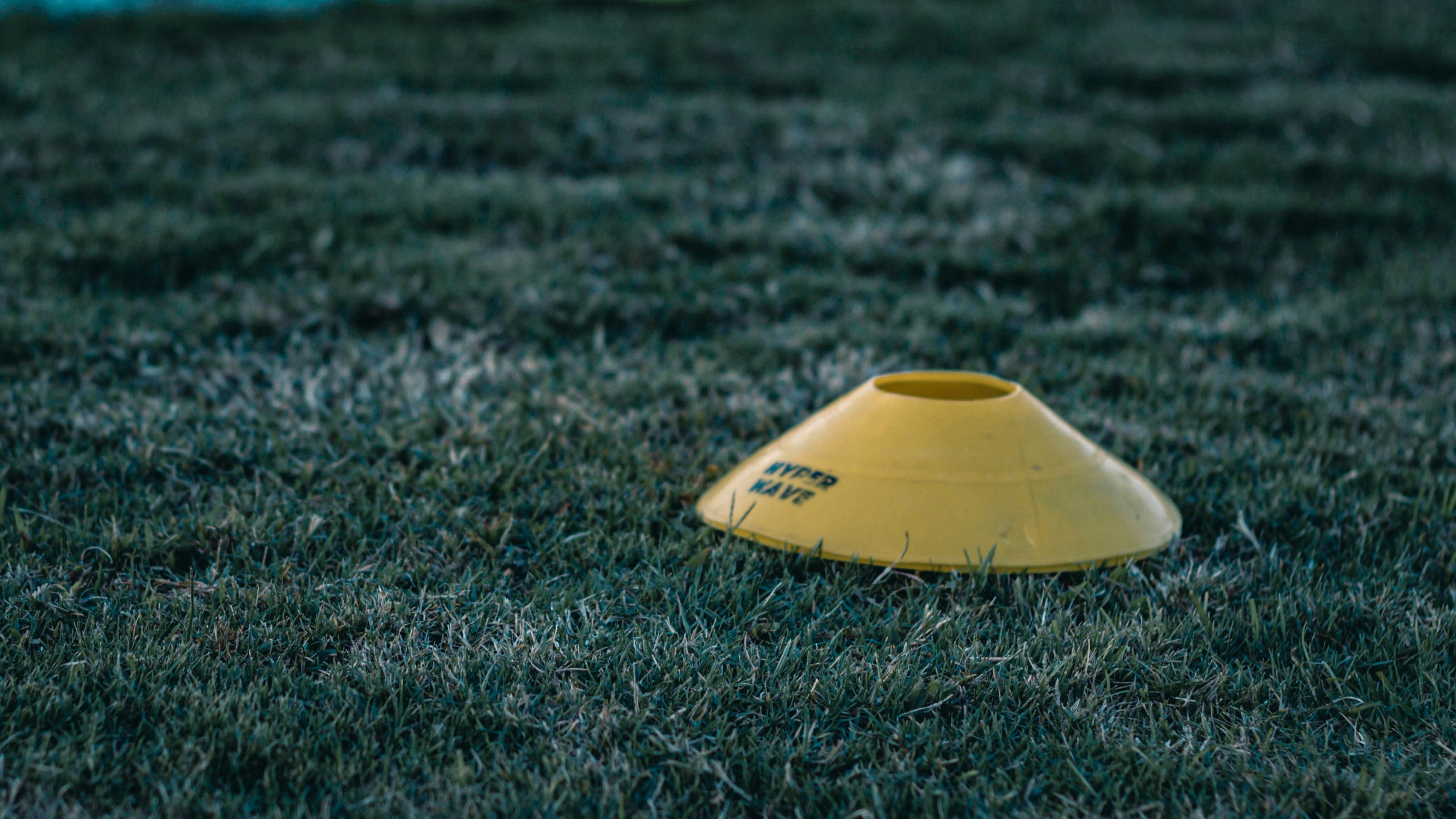 Yellow surface marker resting on a grass field, captured at a wide angle with shallow detail in the foreground; a simple sports-field scene highlighting texture of turf and the object as the focal point.