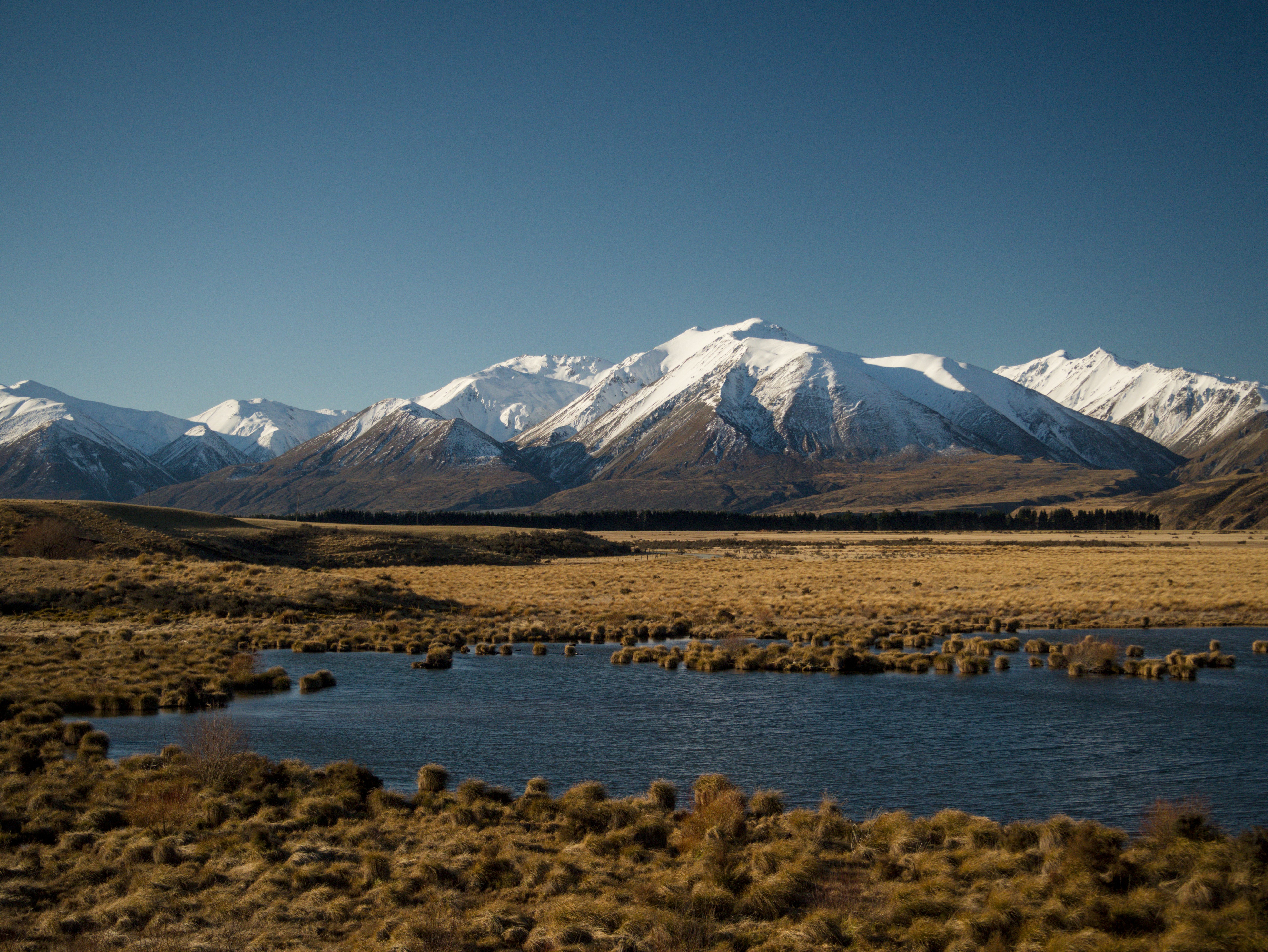 Snowy mountains rise beyond golden heath and a calm waterway, forming a tranquil high-desert scene with distant foothills and a clear blue sky.