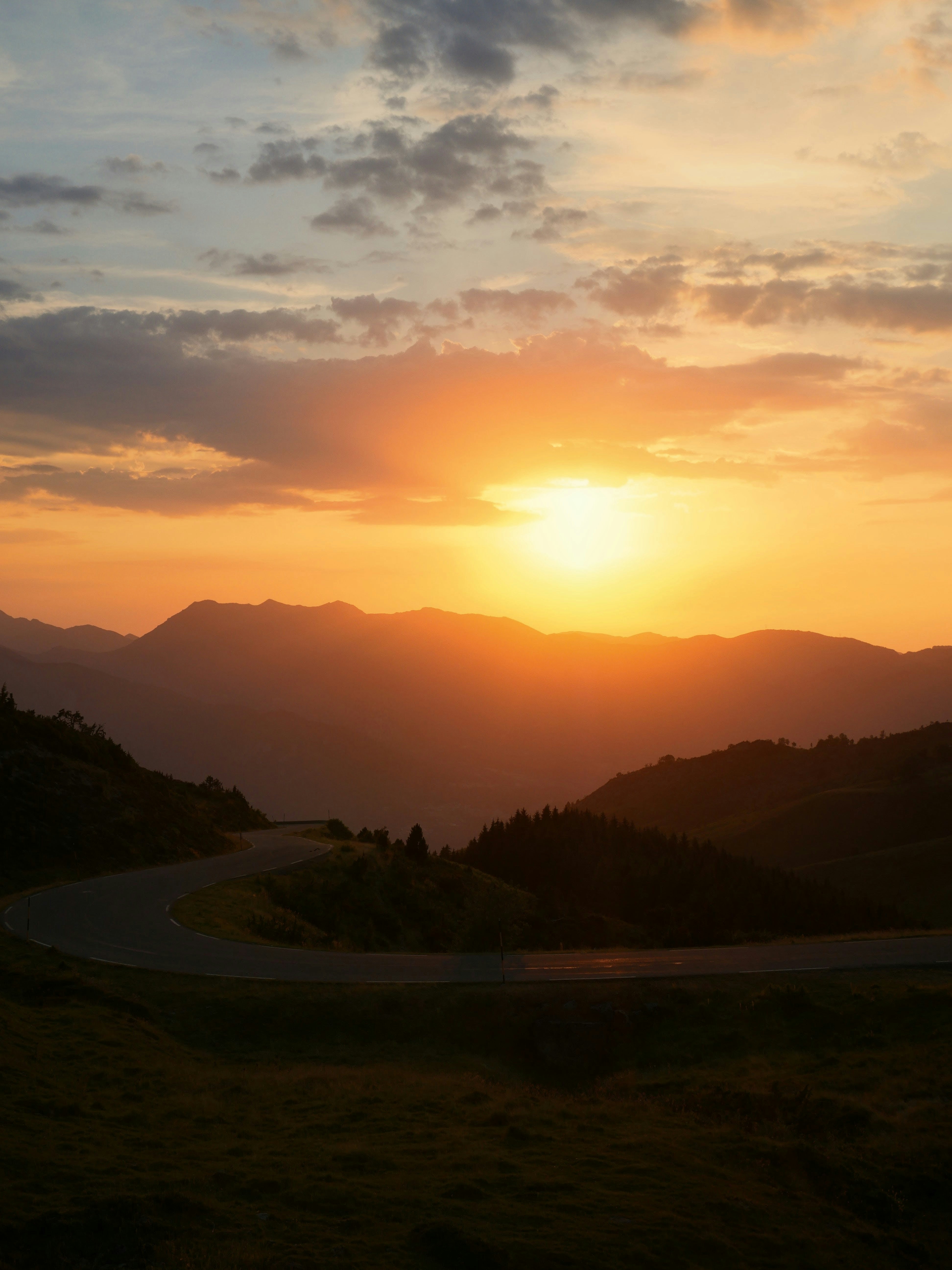 A spectacular sunset over a winding alpine road in Hautacam, Pyrenees, France. The golden sun dips behind layered mountain silhouettes, bathing the sky in vibrant shades of warm orange, yellow, and soft clouds. The empty asphalt road curves through the tranquil landscape, inviting thoughts of evening road trips and serene mountain adventures.