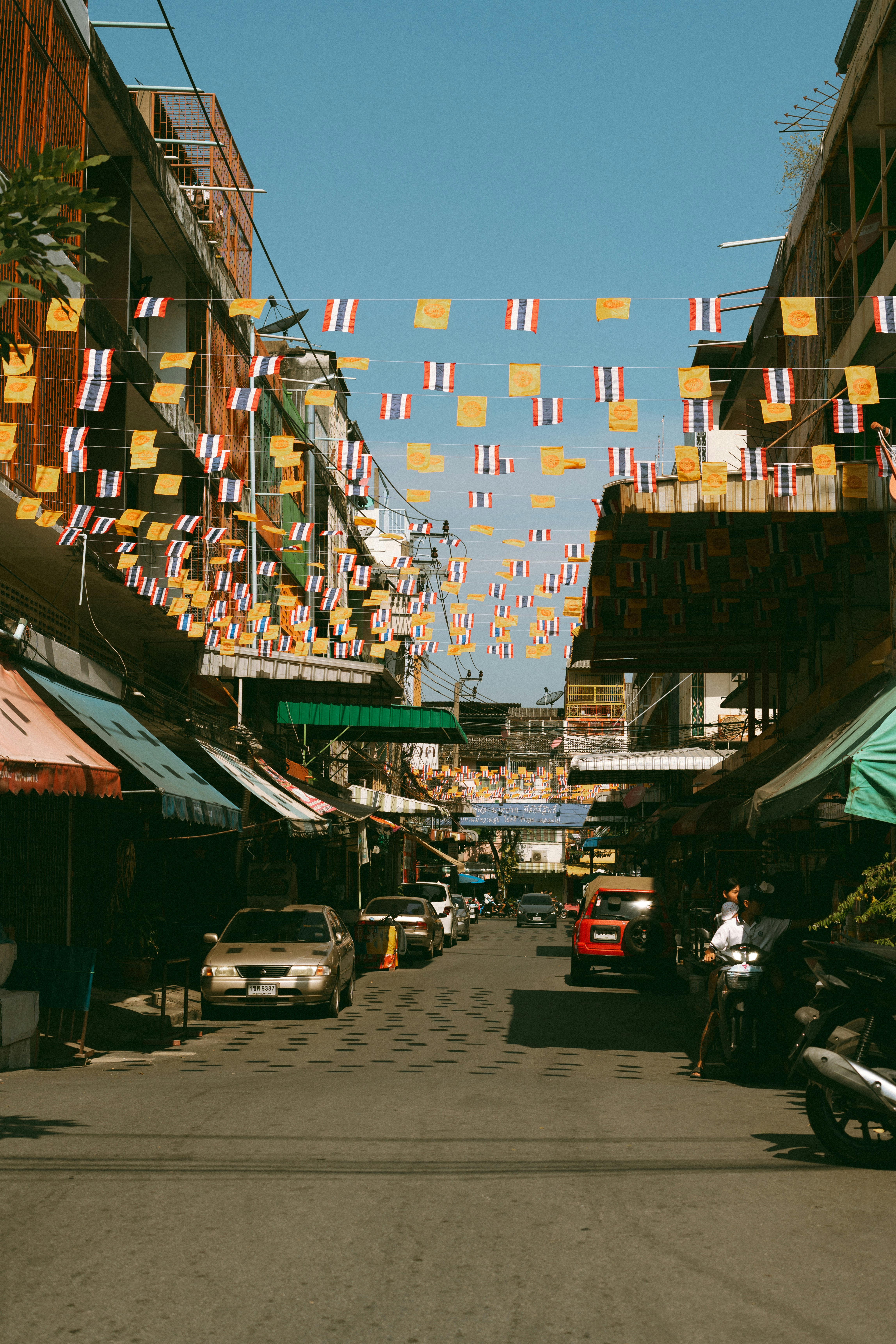Sunlit street market under colorful pennant banners, cars and a scooter line the road while shaded stalls glow with warm hues.