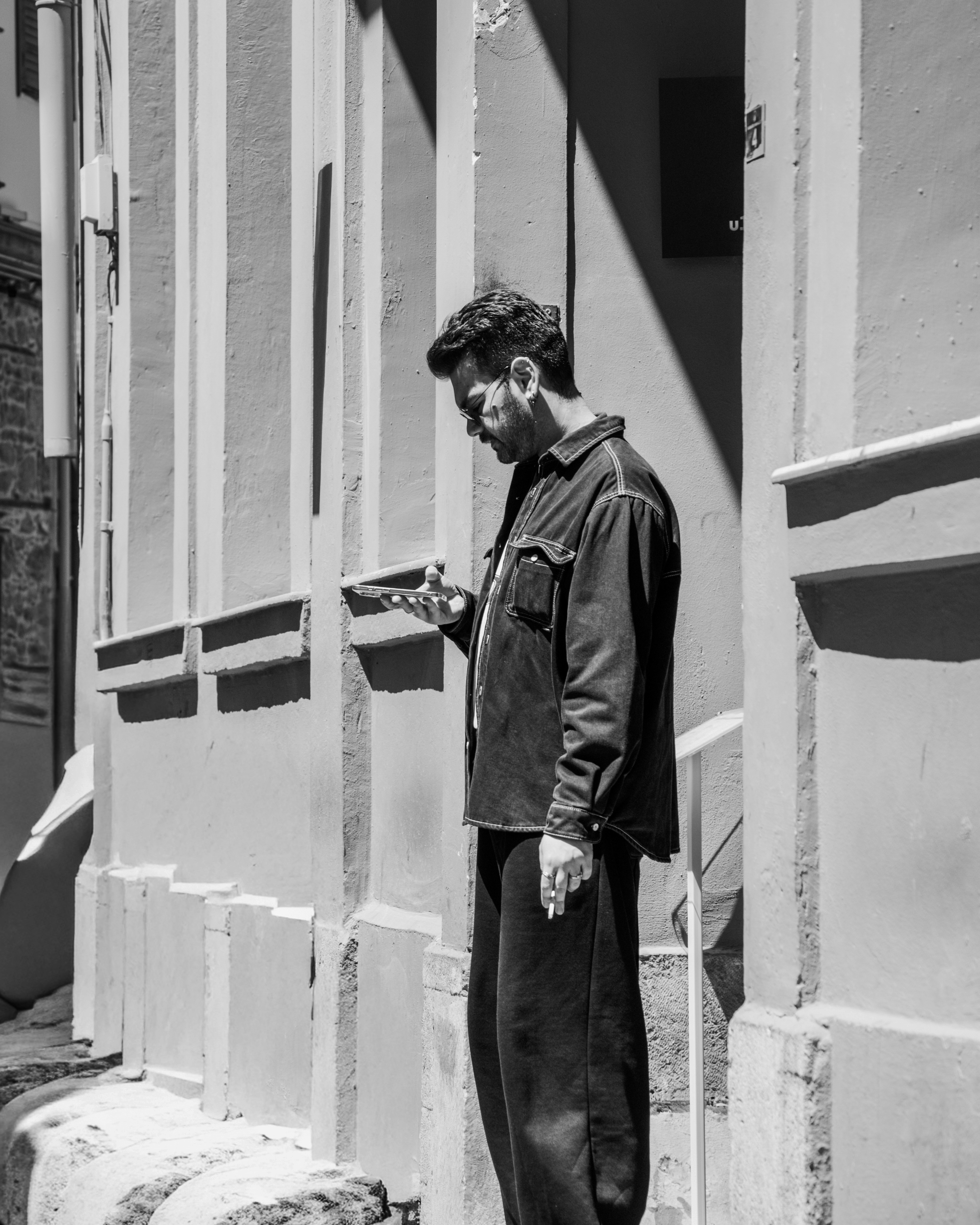 Man with tablet and crutch leaning against sunlit wall in grayscale alley