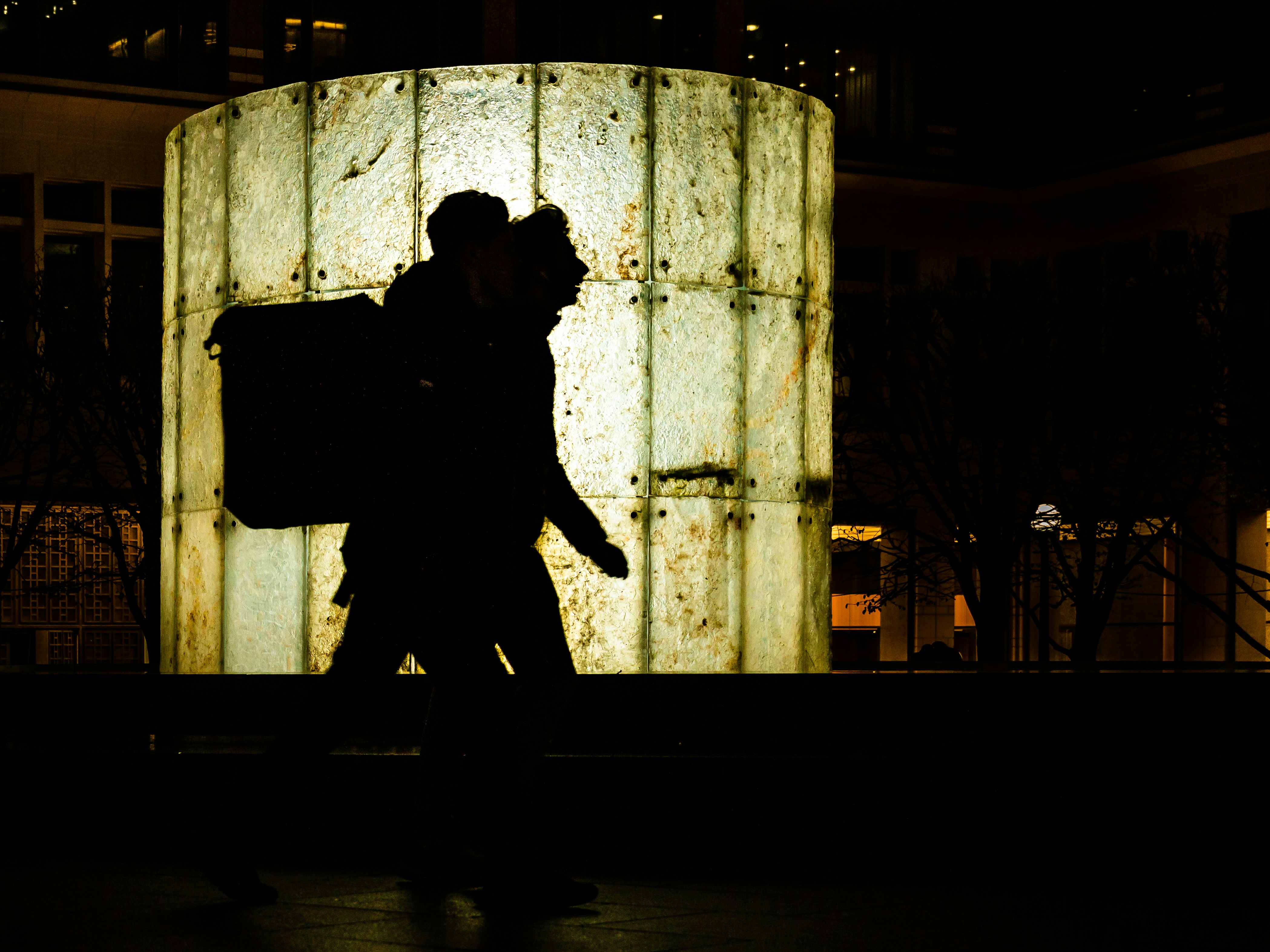 A food delivery courier carrying a backpack walks past an illuminated glass structure at night in London. A moody street photography image highlighting motion, silhouettes, and urban architecture.