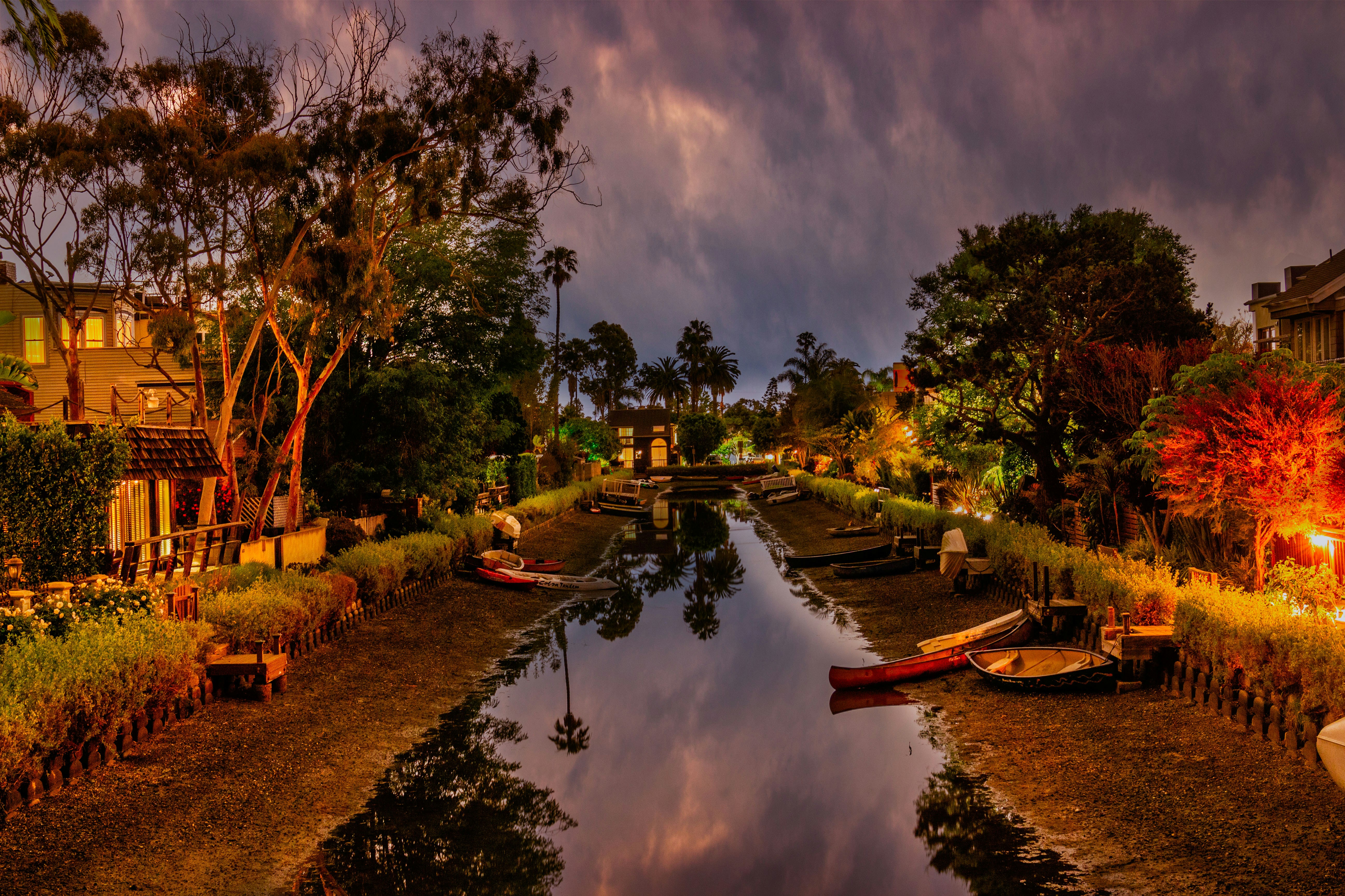 Twilight canal scene with calm water reflecting a pink-orange sky; boats line the banks of a small town, trees and houses illuminated by warm street lighting.