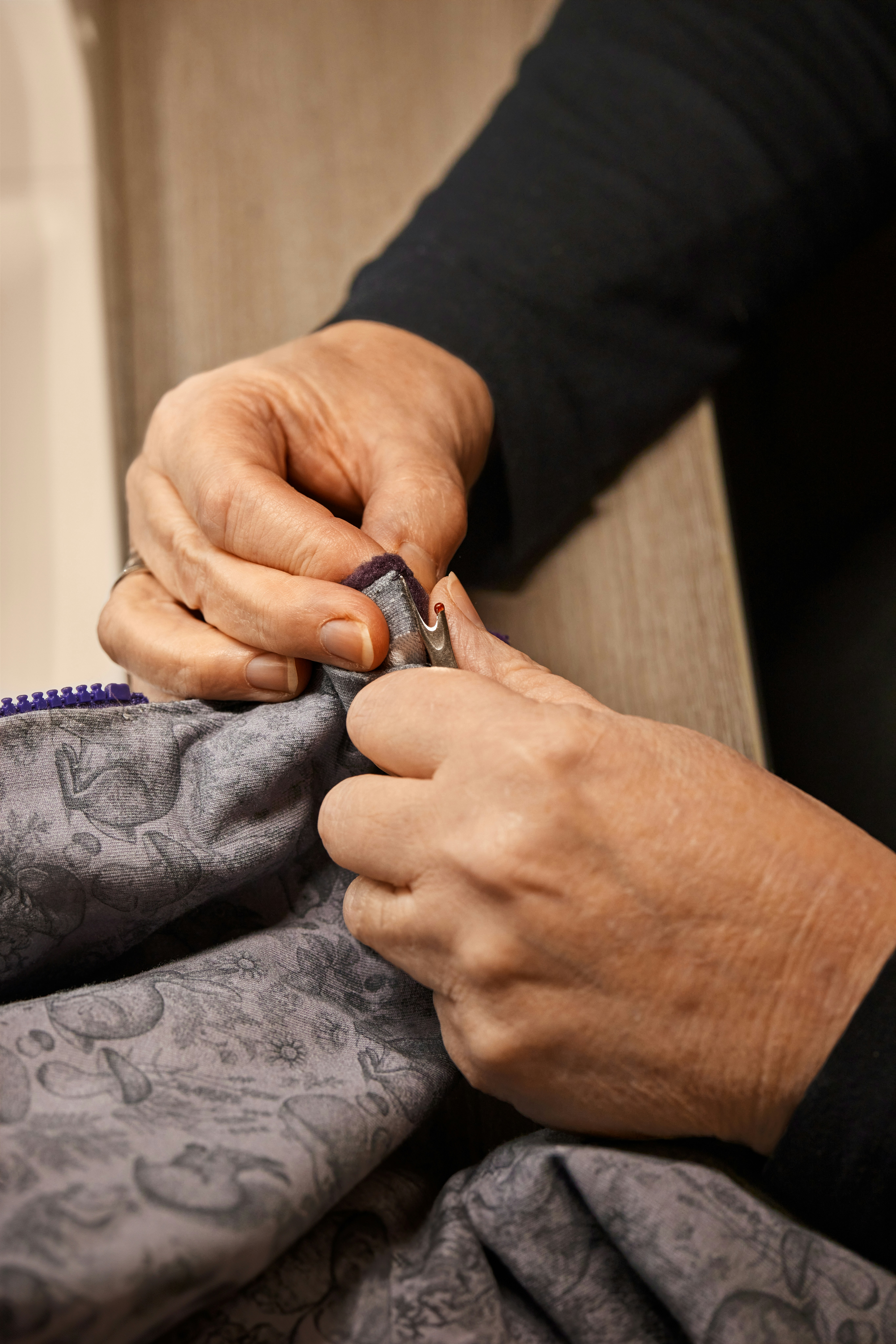 Close-up of hands weaving a zipper into a gray patterned fabric, showing careful stitching and fabric manipulation.
