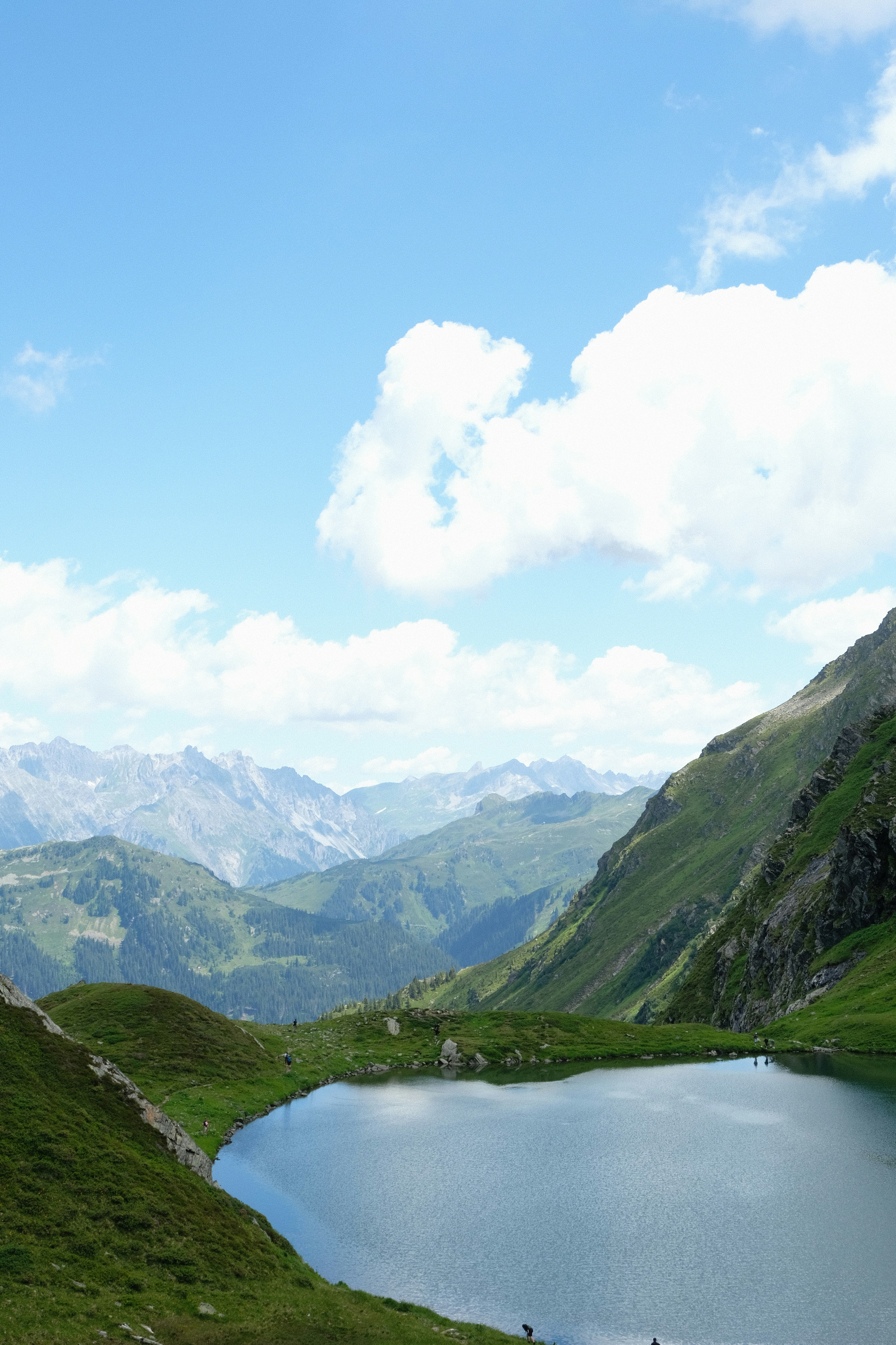 Bright alpine lake nestles between green slopes under a blue sky with scattered clouds, framed by rugged mountain ridges.